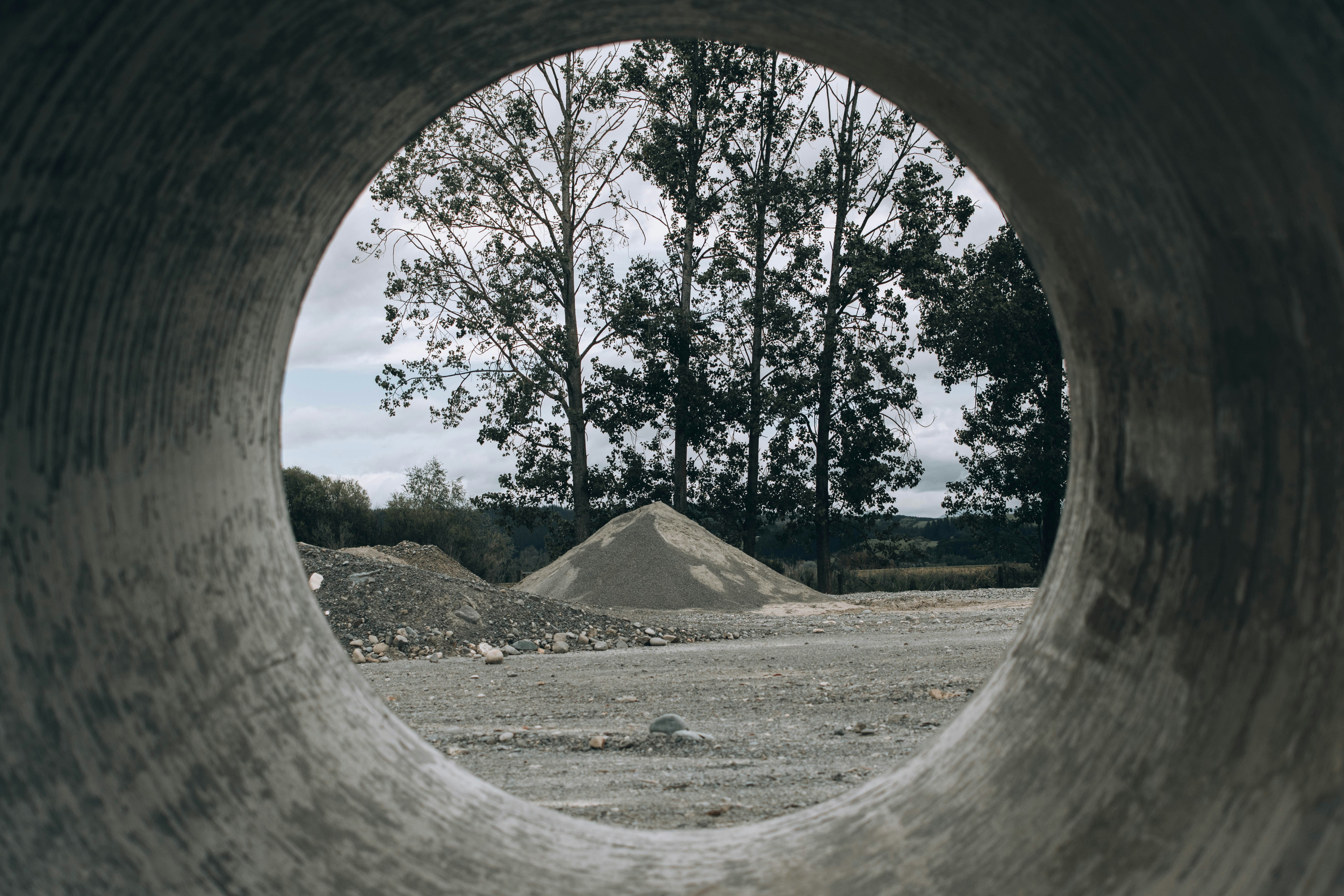 View of trees framed by a concrete pipe, with a gravel mound in the background. The juxtaposition of industrial and natural elements creates a unique composition.