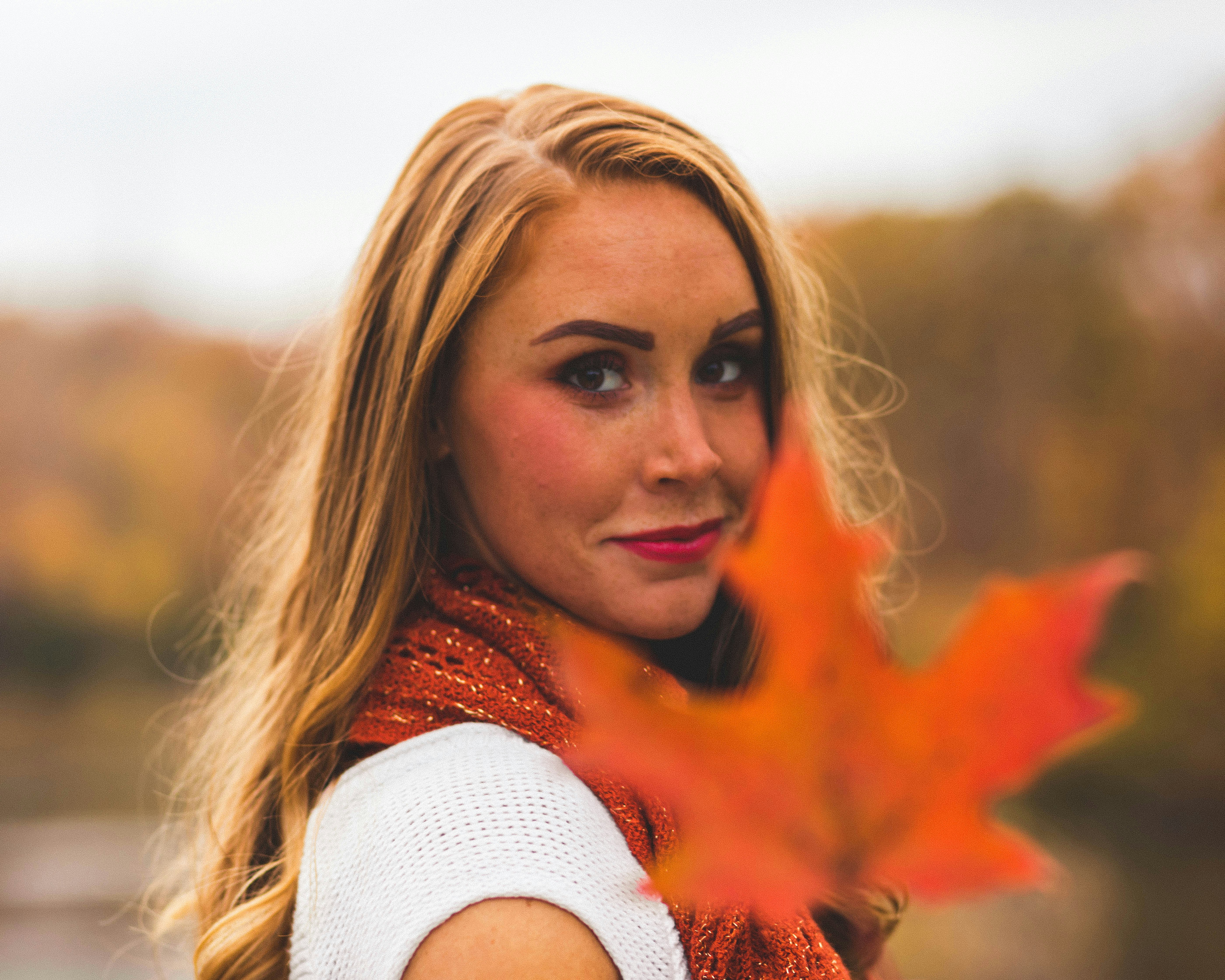 Young woman with long hair and a vibrant scarf gazes over her shoulder, holding a colorful autumn leaf in the foreground.