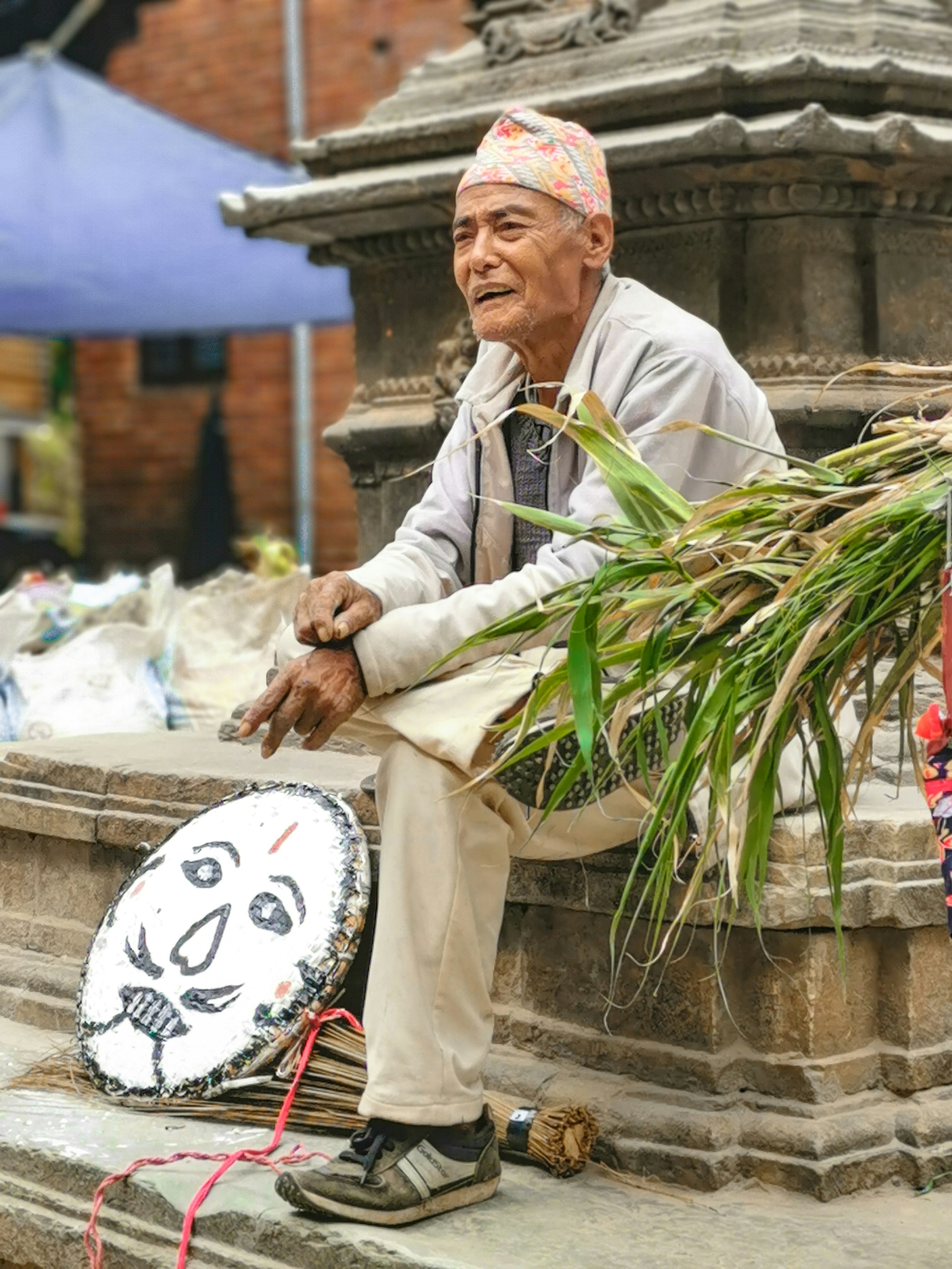 Elderly man seated beside a decorative mask, embodying the essence of local culture and tradition. The scene captures a moment of reflection amidst a vibrant marketplace.