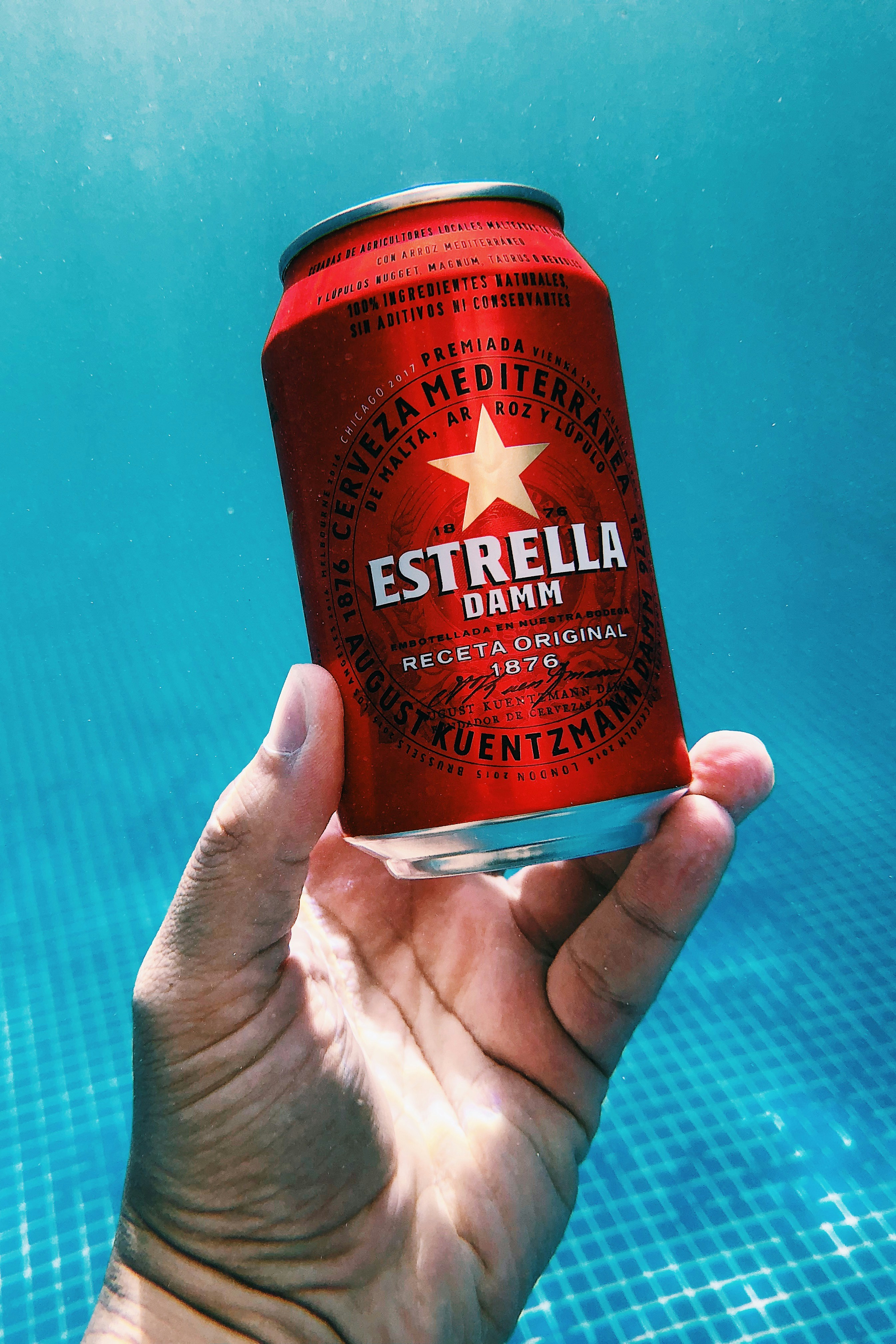 Hand holding a vibrant red Estrella Damm beer can submerged in clear blue water, showcasing its striking design and refreshing appeal.
