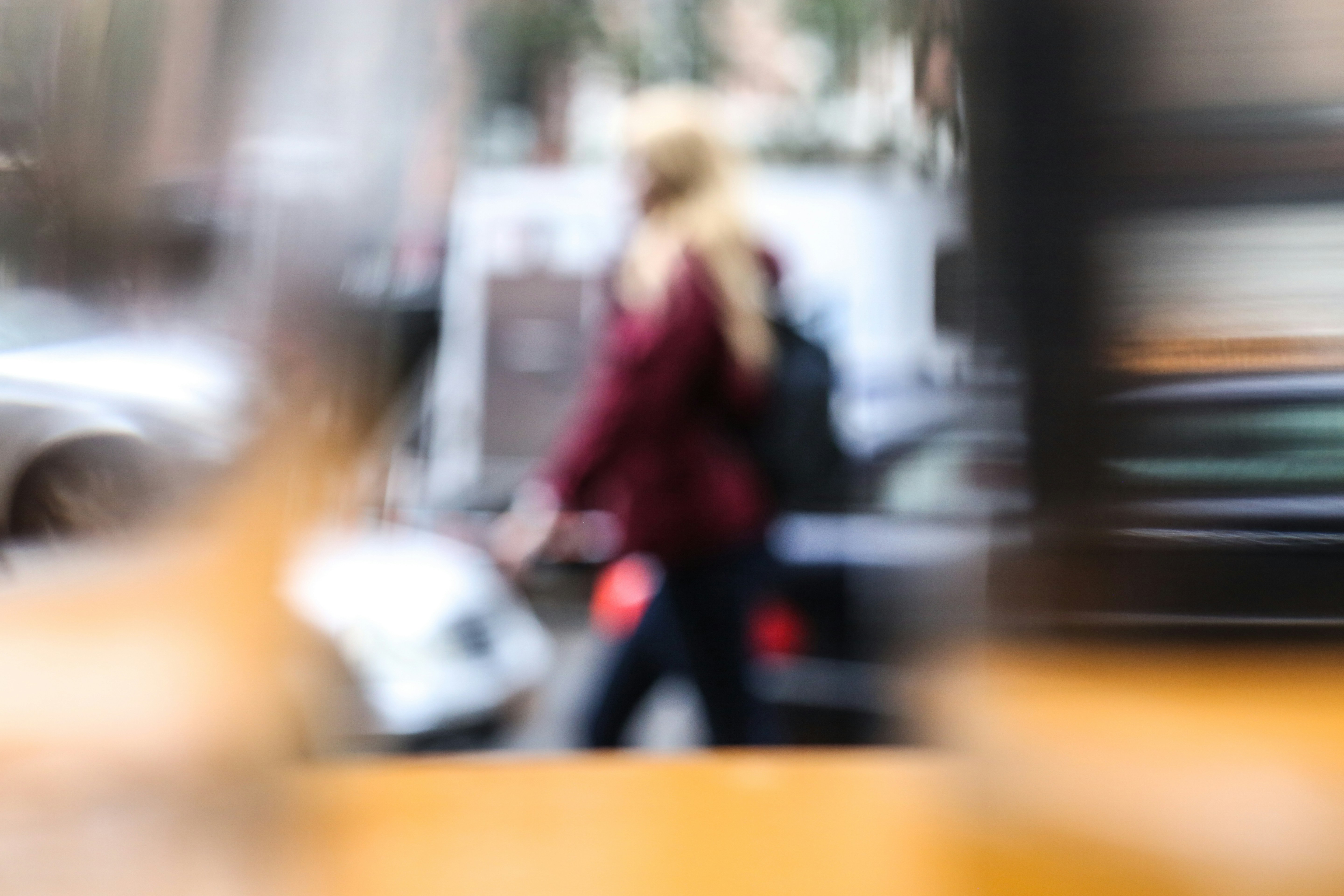 Blurred scene of a person walking past parked cars, viewed through a glass surface. The motion conveys a sense of urban life.