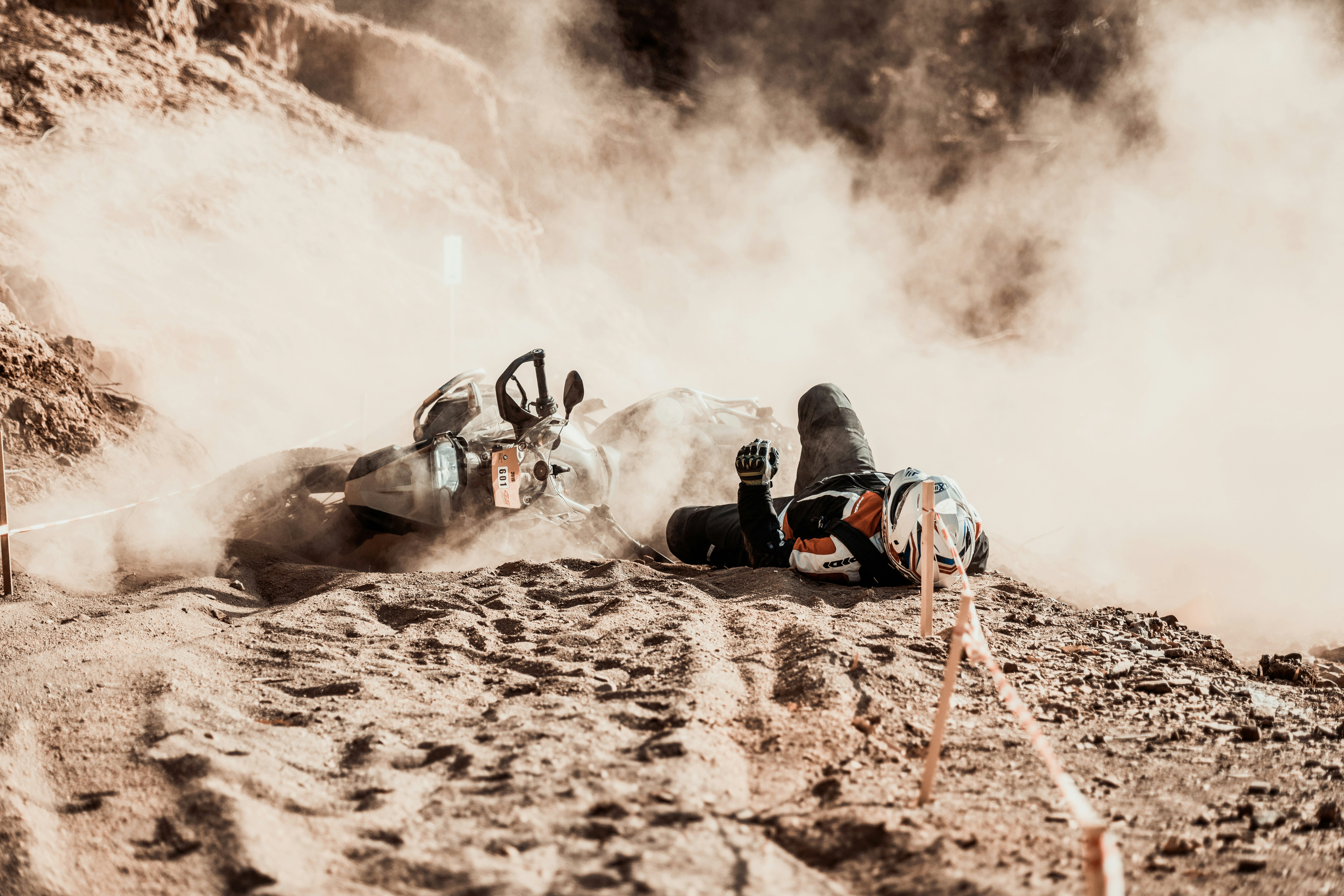 Motorcycle rider sprawled on sandy terrain amidst a cloud of dust after a fall during a race.