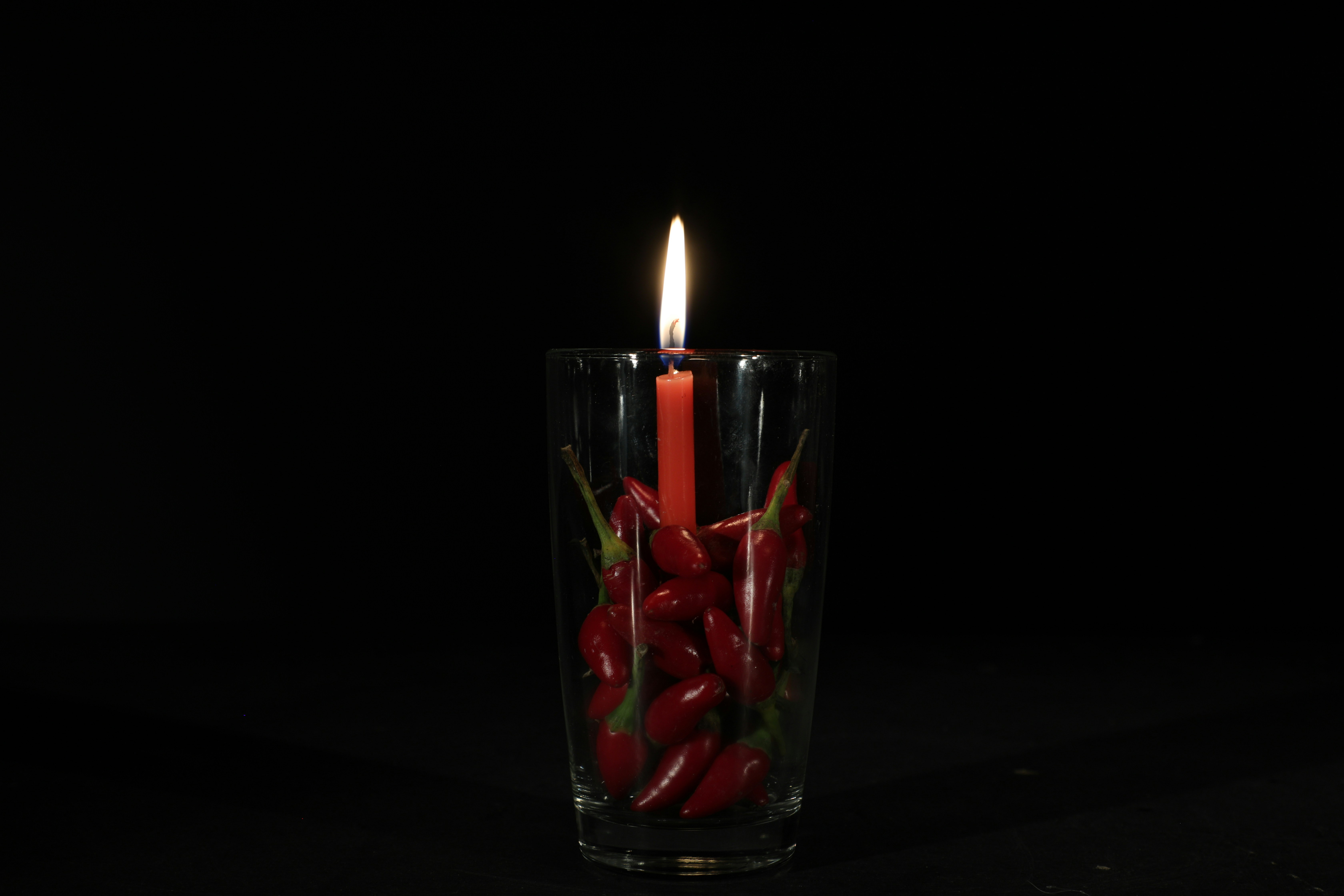 A vibrant red candle stands tall amidst a cluster of red chili peppers in a clear glass, illuminated against a dark backdrop.