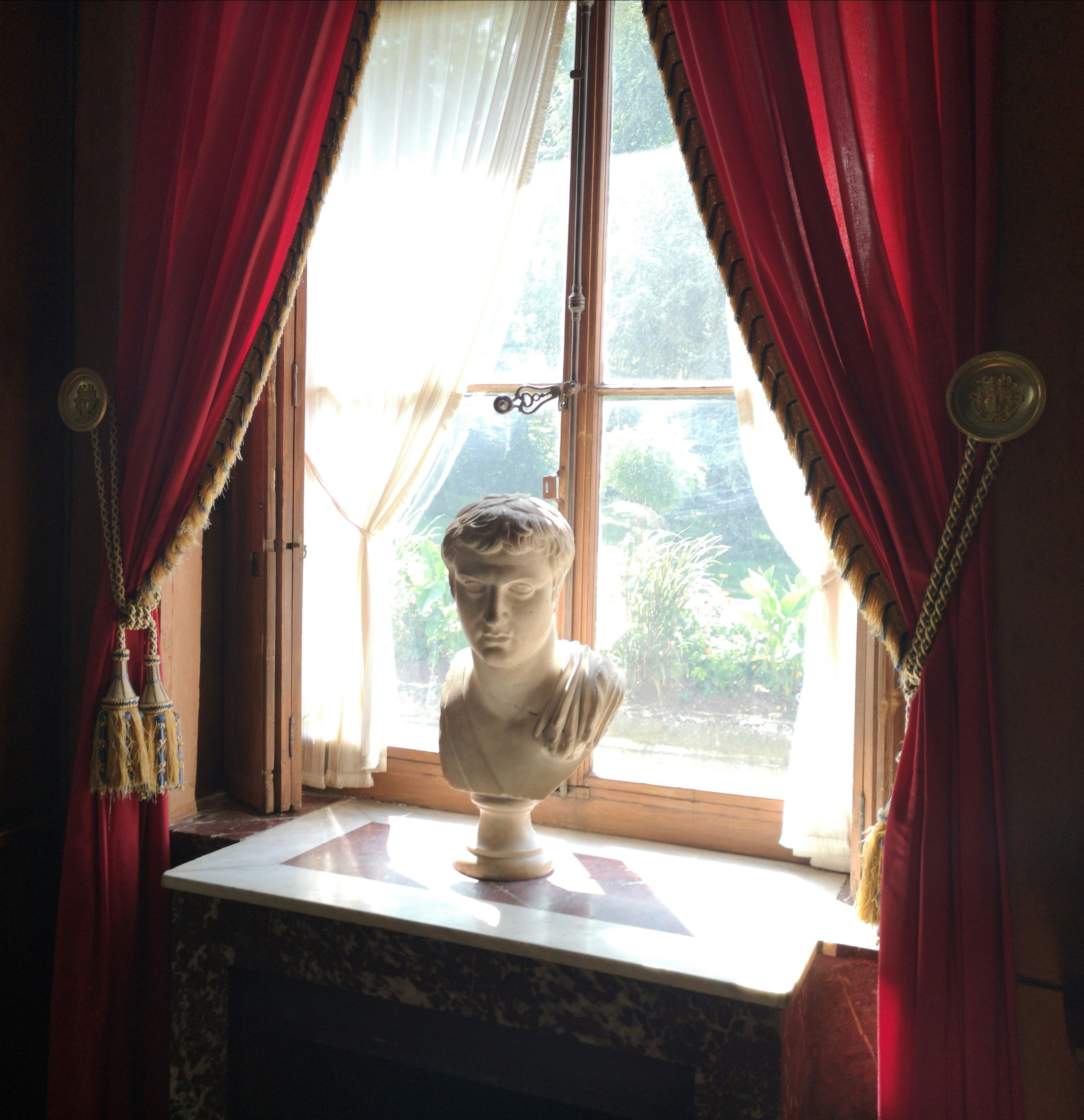 Classical bust sculpture positioned on a marble pedestal, framed by rich red curtains and illuminated by natural light from a window.