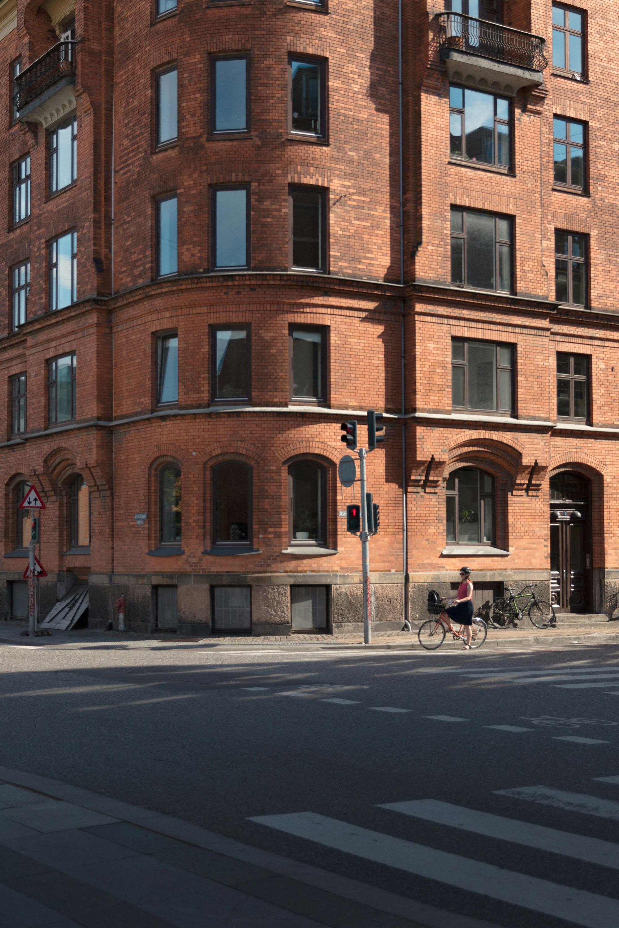 A brick building with large windows at a city intersection, featuring a cyclist waiting at a traffic light. The scene captures the essence of urban living.