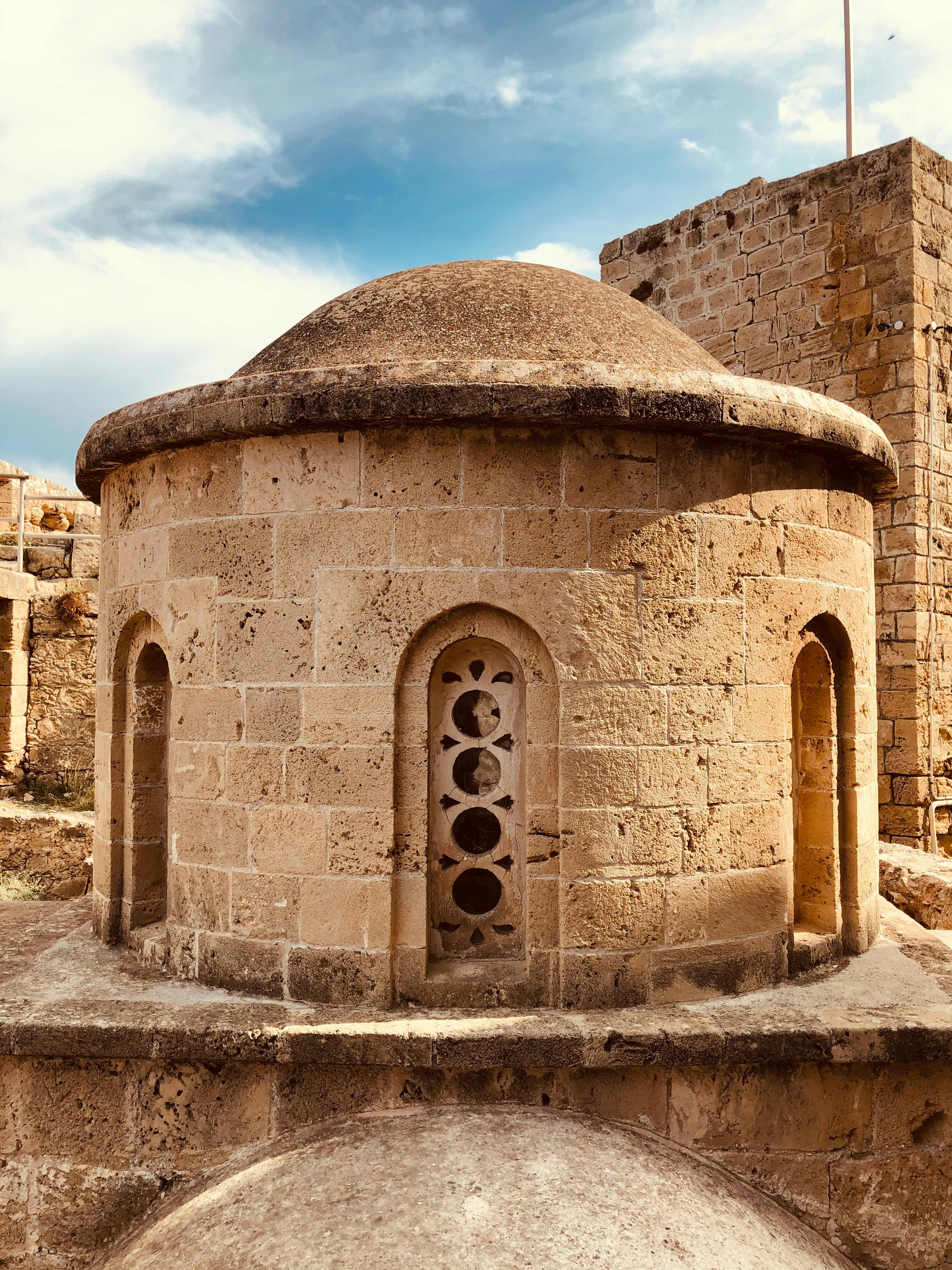 A weathered stone dome structure adorned with arched windows, set against a backdrop of blue skies and rugged architecture. The scene evokes a sense of history and tranquility.