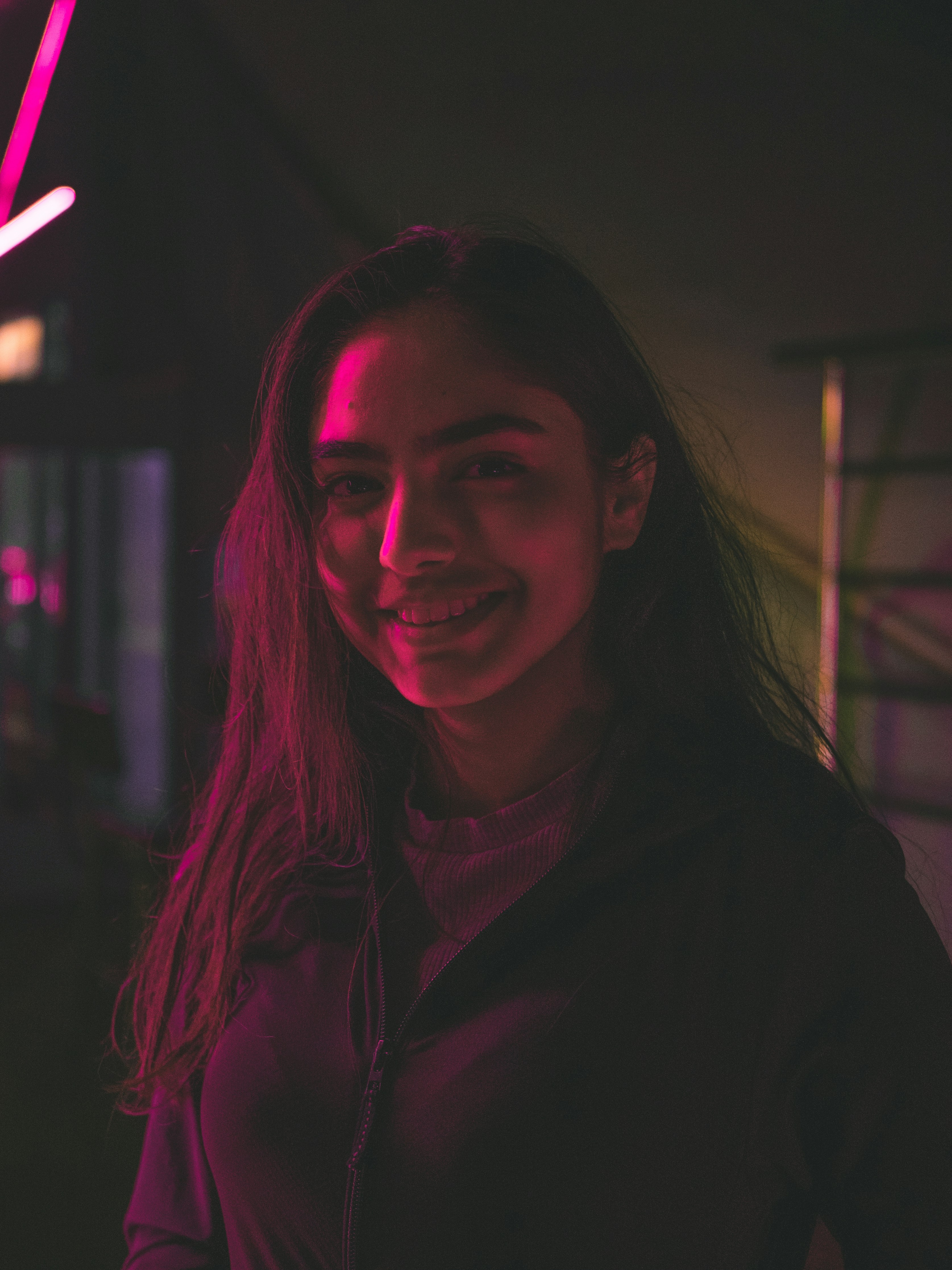 Portrait of a smiling woman bathed in magenta neon light in a dark indoor space, creating a moody, color-rich atmosphere.