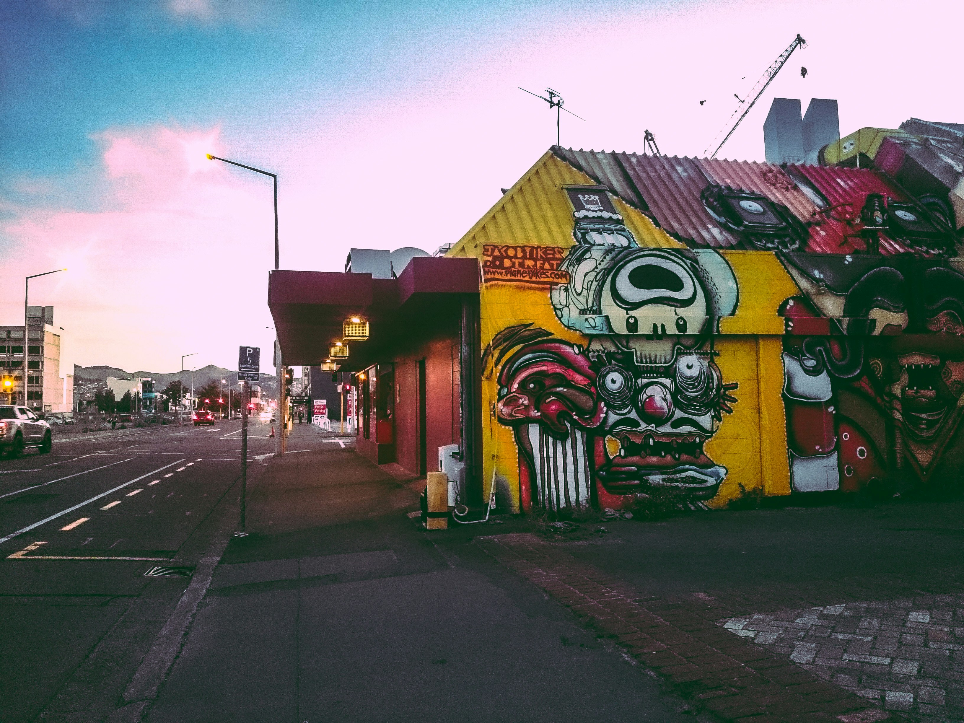 Street art mural on a yellow building under a dusky sky, with city street and traffic in the background.