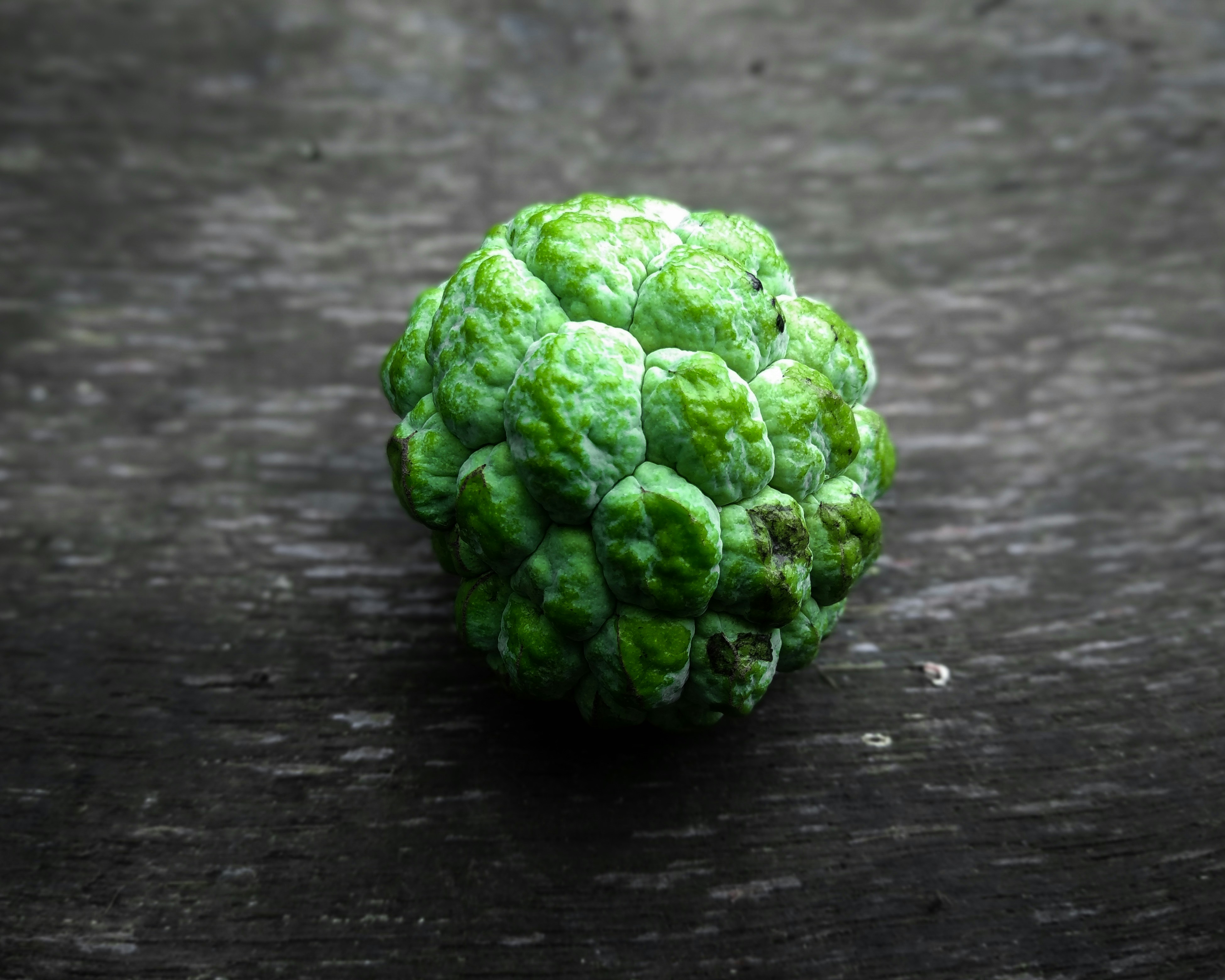 A single green sugar-apple rests on dark, weathered wood, its segmented surface rendered in sharp relief.