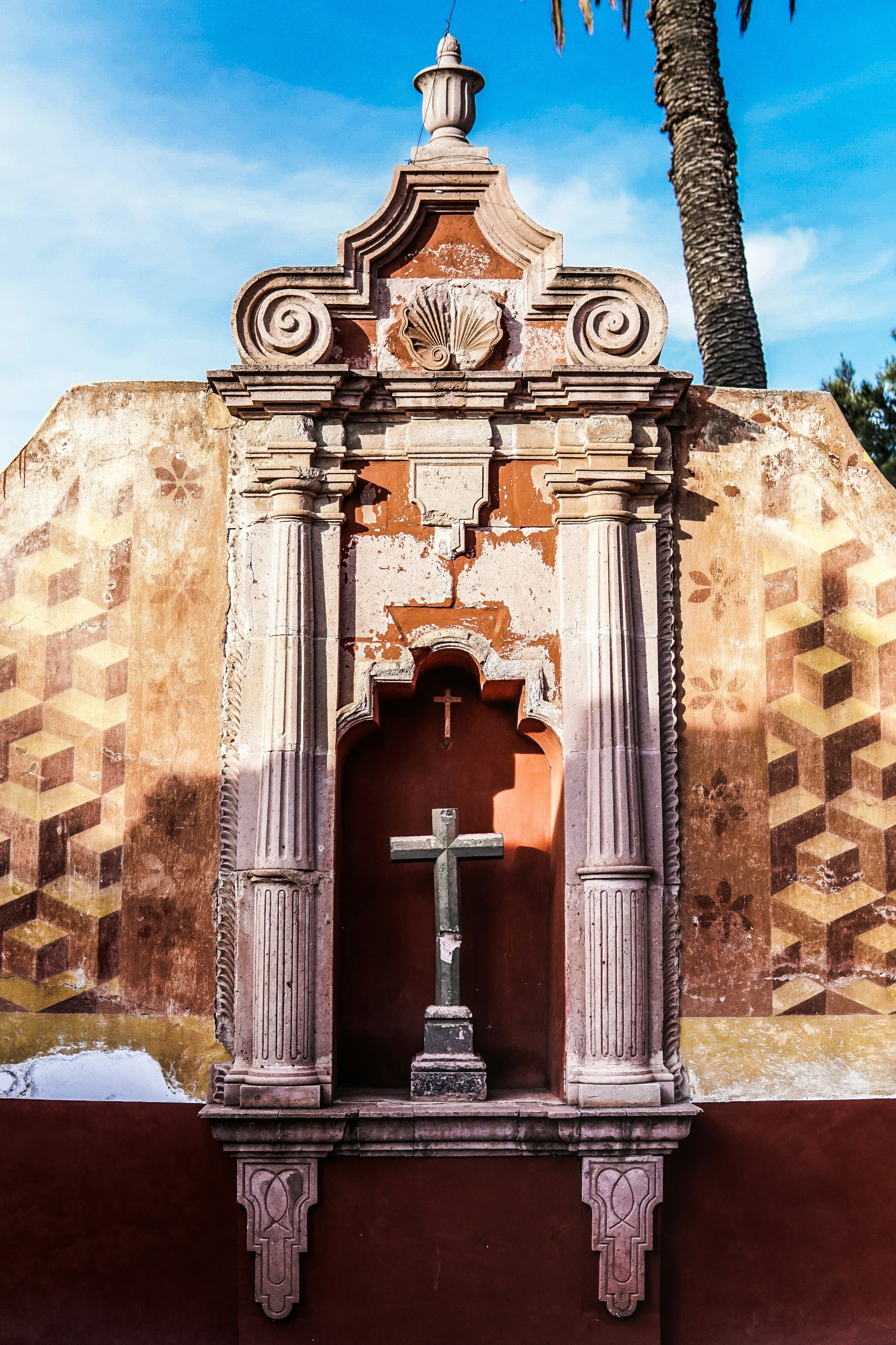 Ornate stone alcove with a cross against a patterned wall under a blue sky.