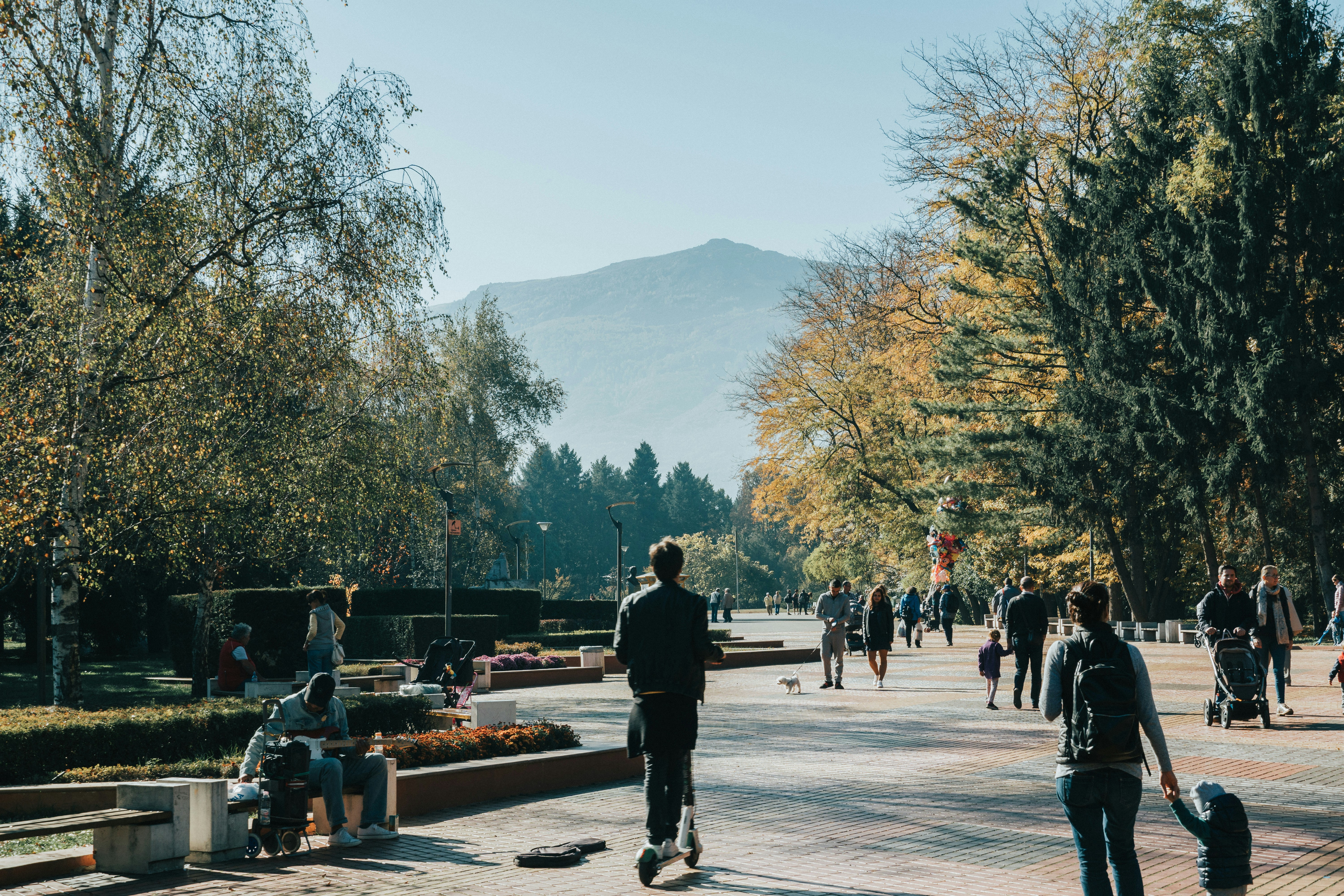 People walking and riding scooters along a sunlit path with autumn trees and a mountain in the background.