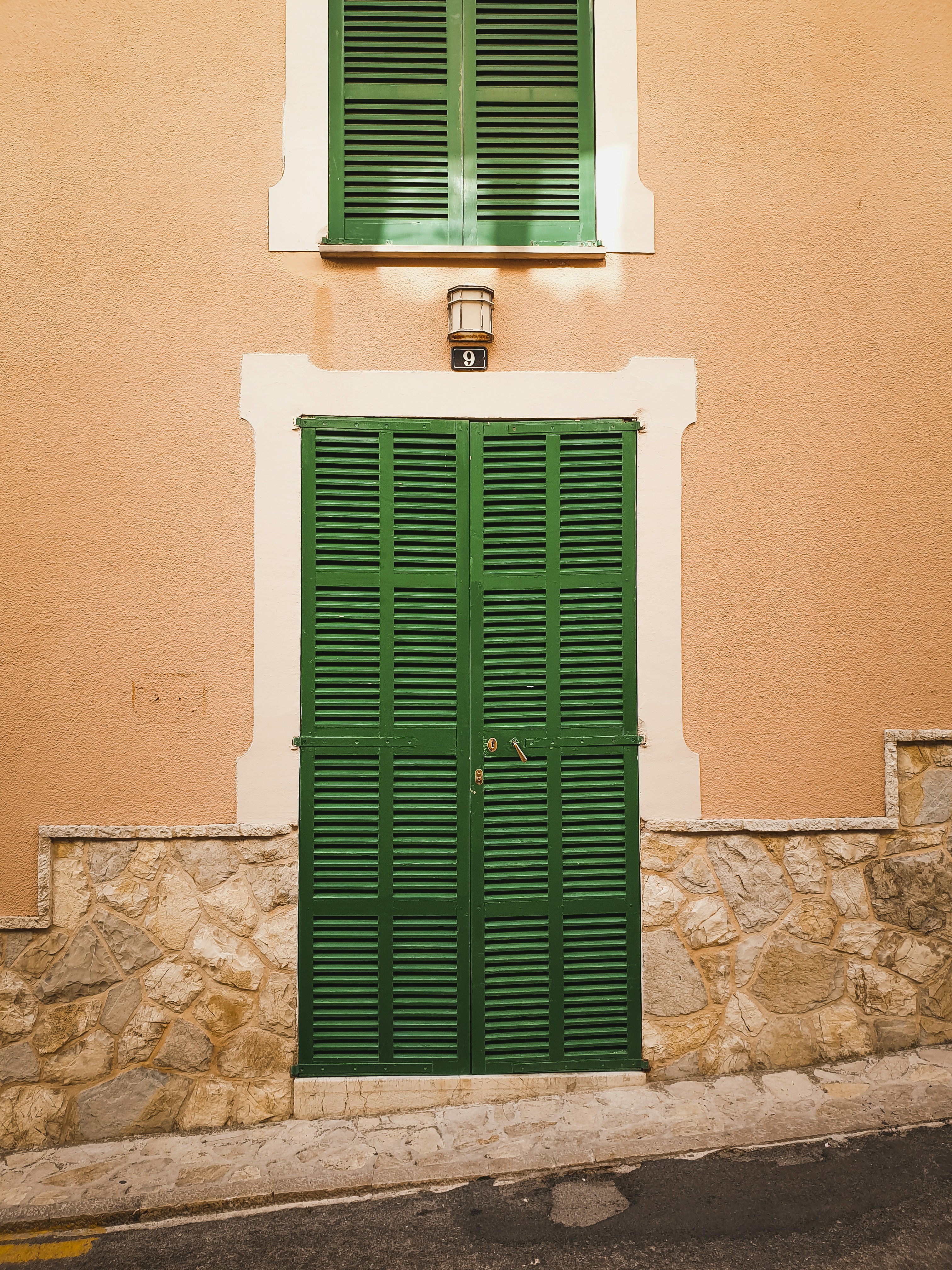 Green louvered shutters frame a peach stucco facade with a stone base, creating a centered, sunlit street scene.