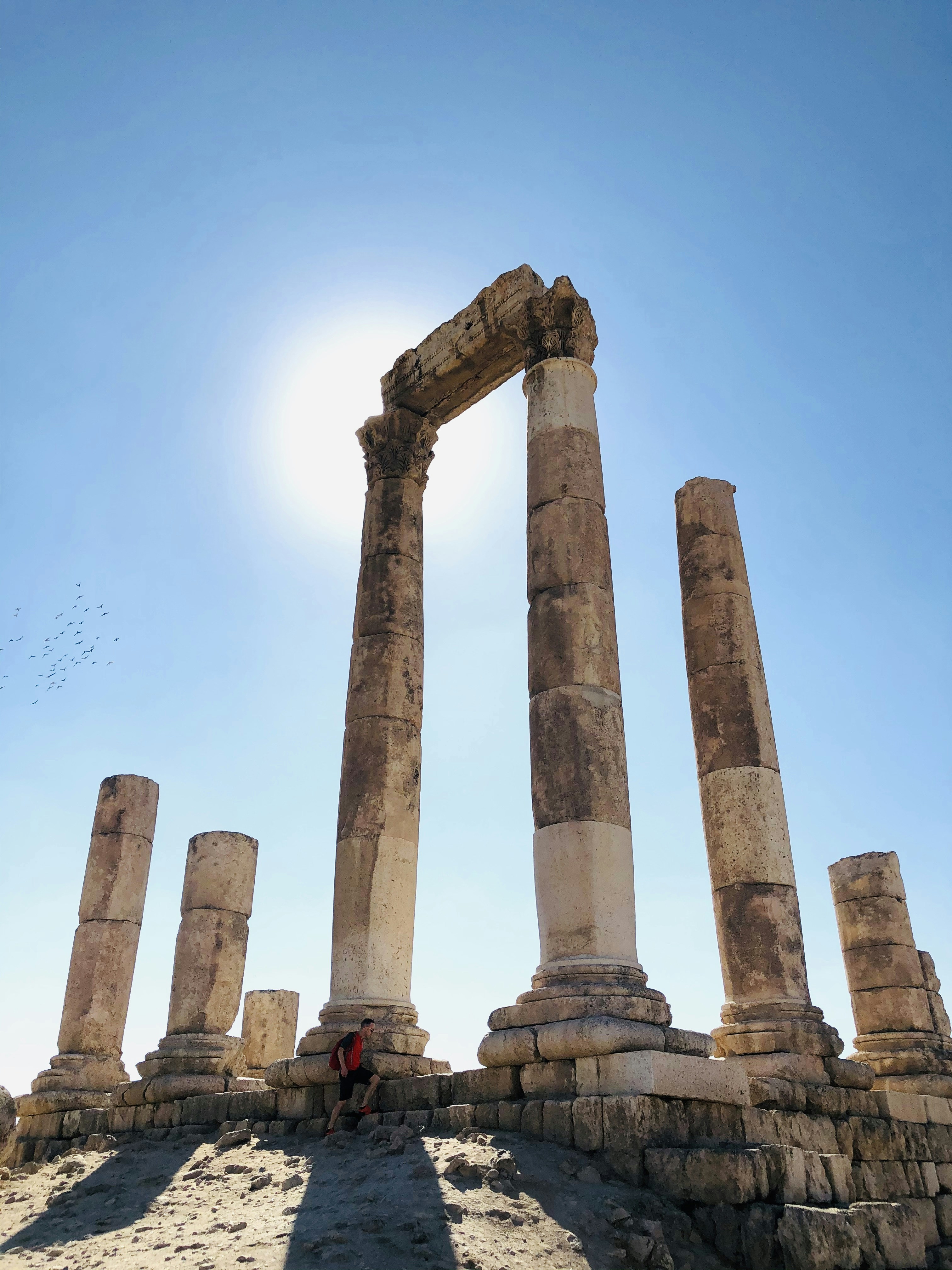 Ancient stone columns rise against a clear blue sky, with a person seated at the base, evoking a sense of history and scale. Sunlight creates a halo effect around the top structure.