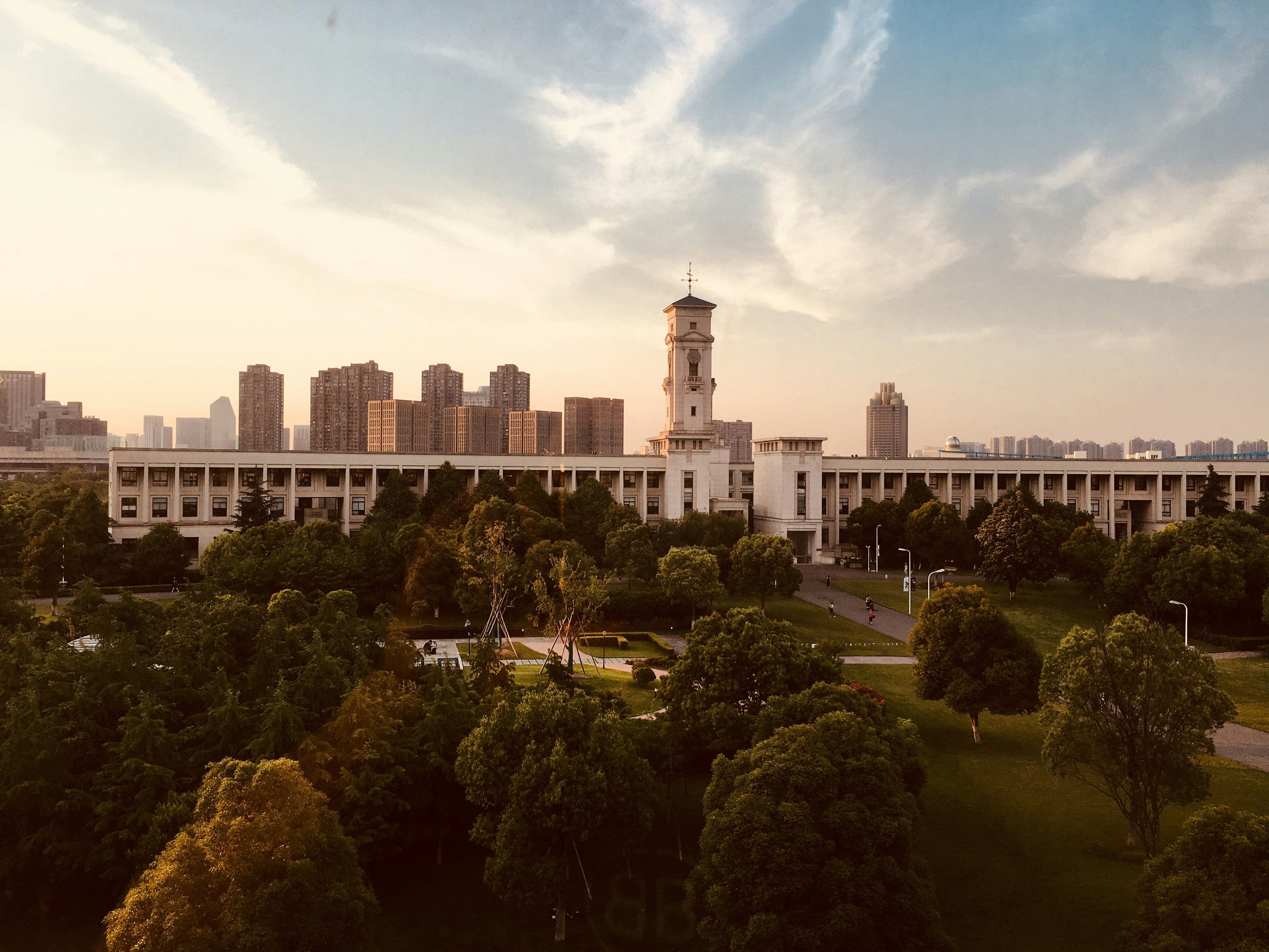 Clock tower rises above lush trees with a city skyline in the background under a dynamic sky.