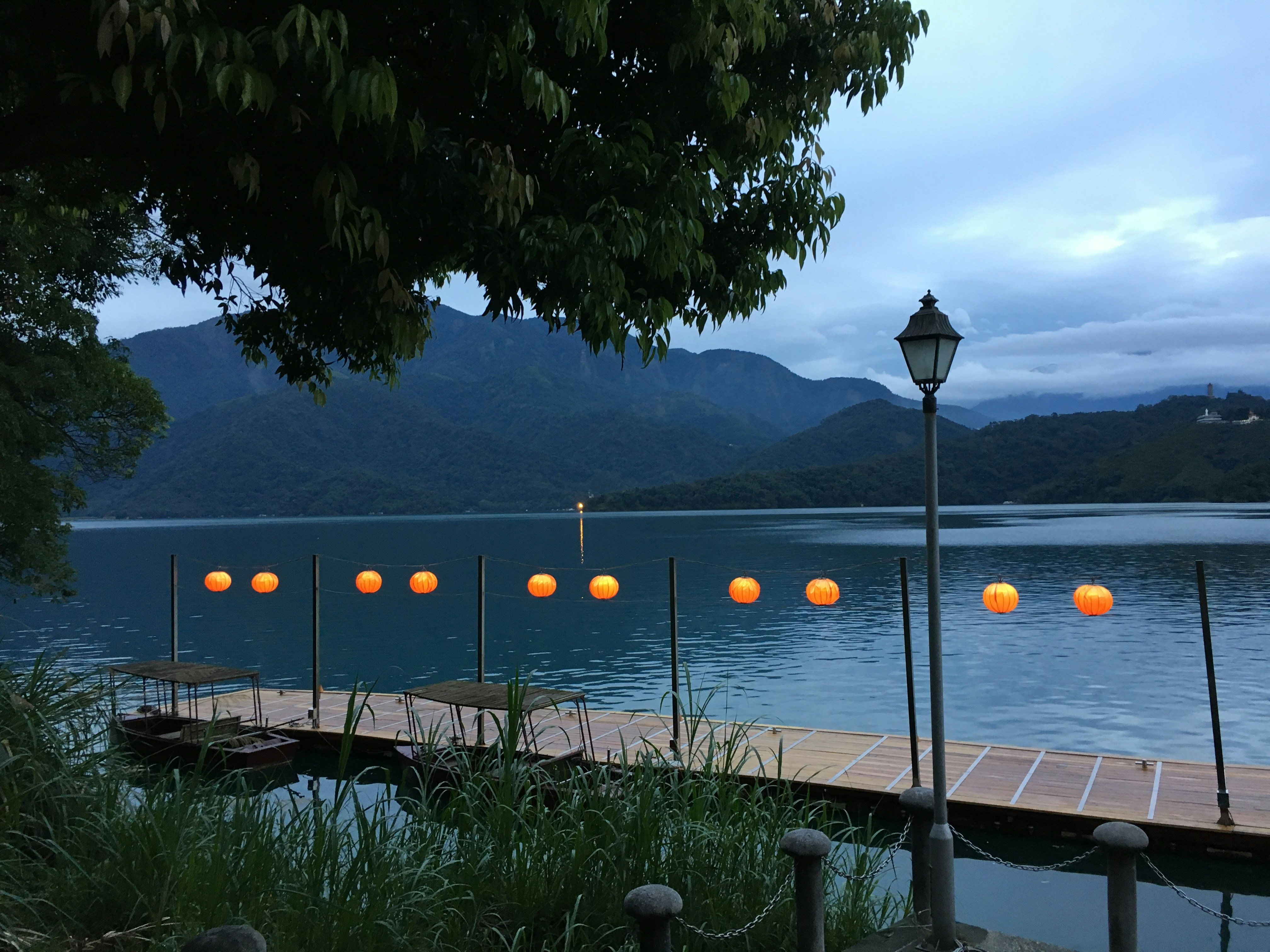 Serene lakeside pier with glowing lamps against a backdrop of mountains under a cloudy evening sky.