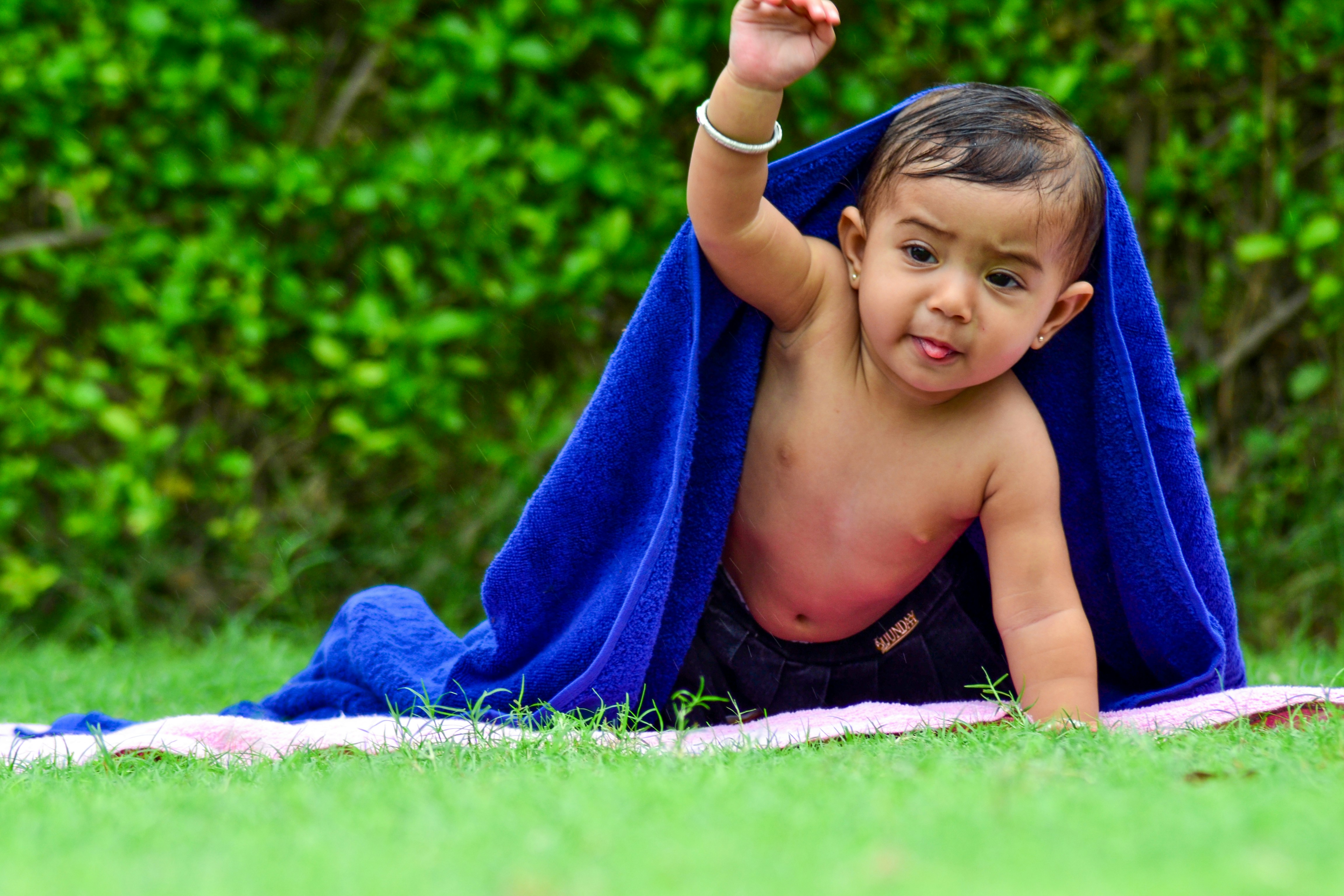 A toddler playfully peeks out from under a blue towel, surrounded by lush green grass, showcasing innocence and curiosity.