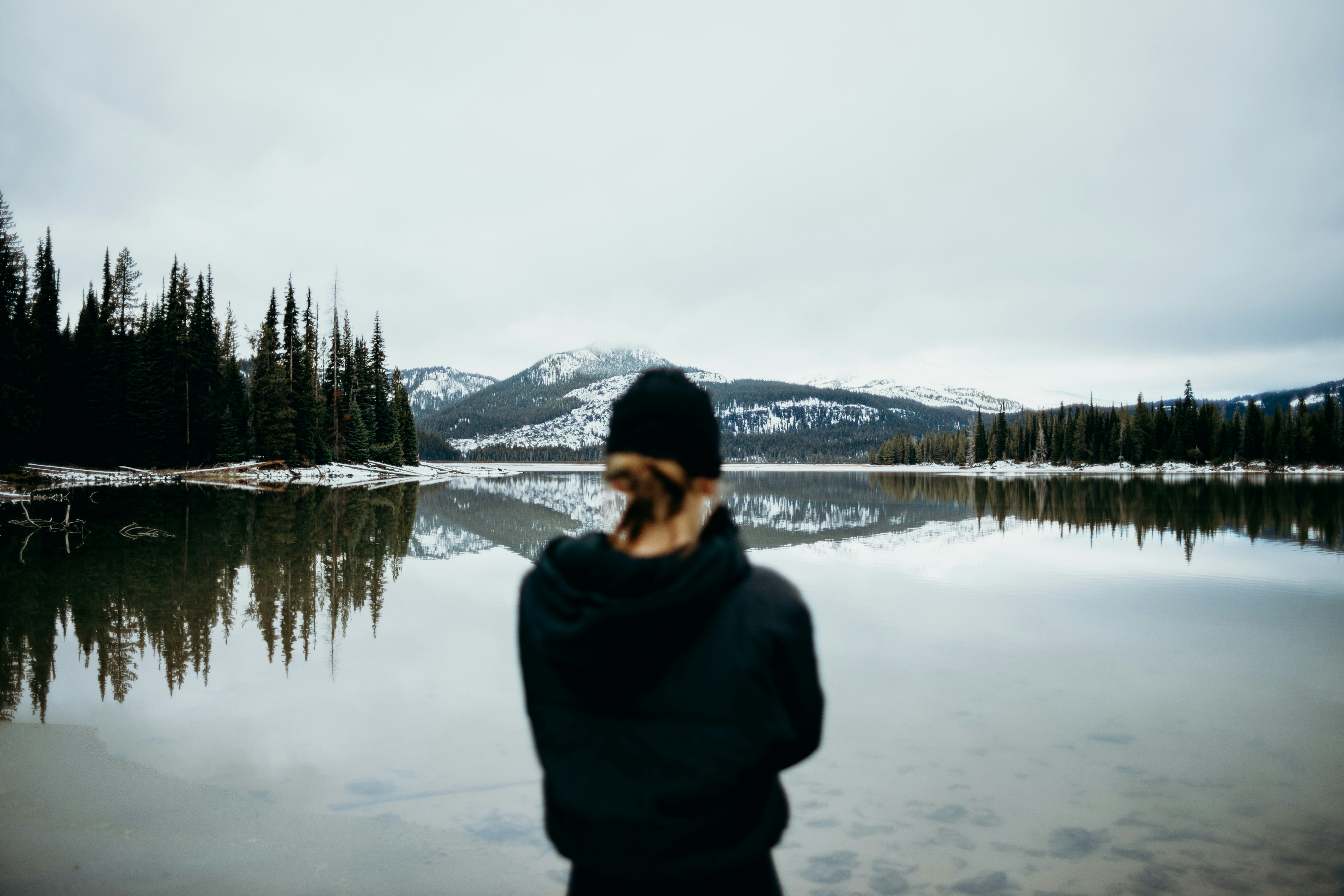 Person in a beanie stands before a tranquil lake reflecting snow-capped mountains and evergreen trees.