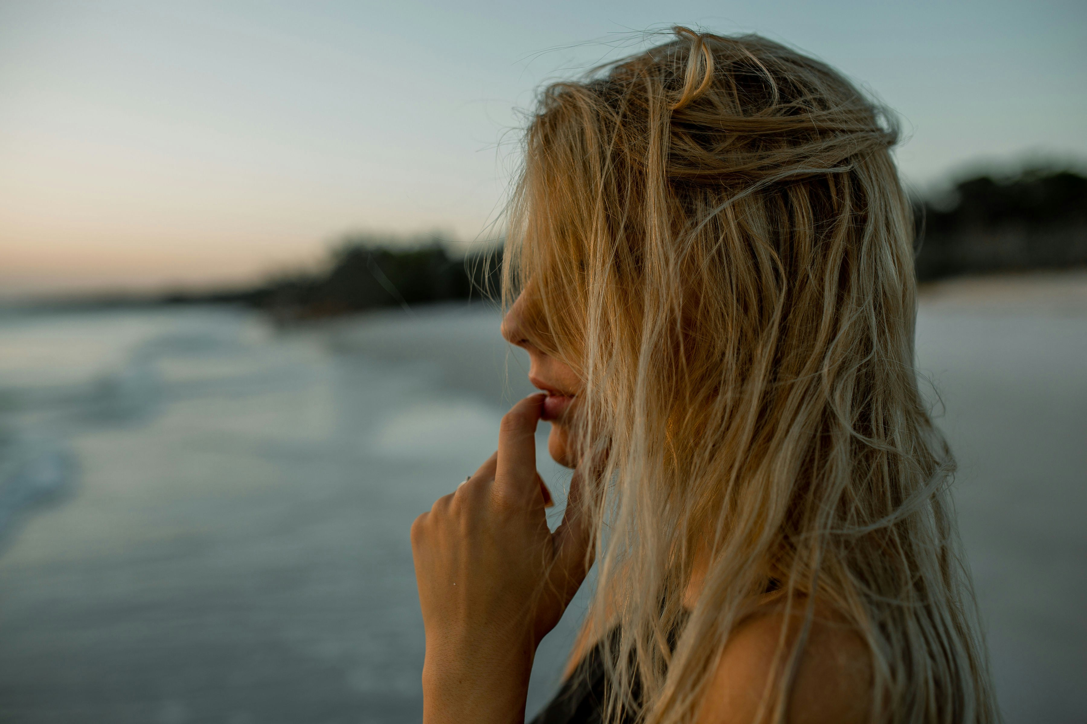 A contemplative young woman stands by the shore, lost in thought as the waves gently lap at the sand. Her flowing hair catches the soft light of dusk.