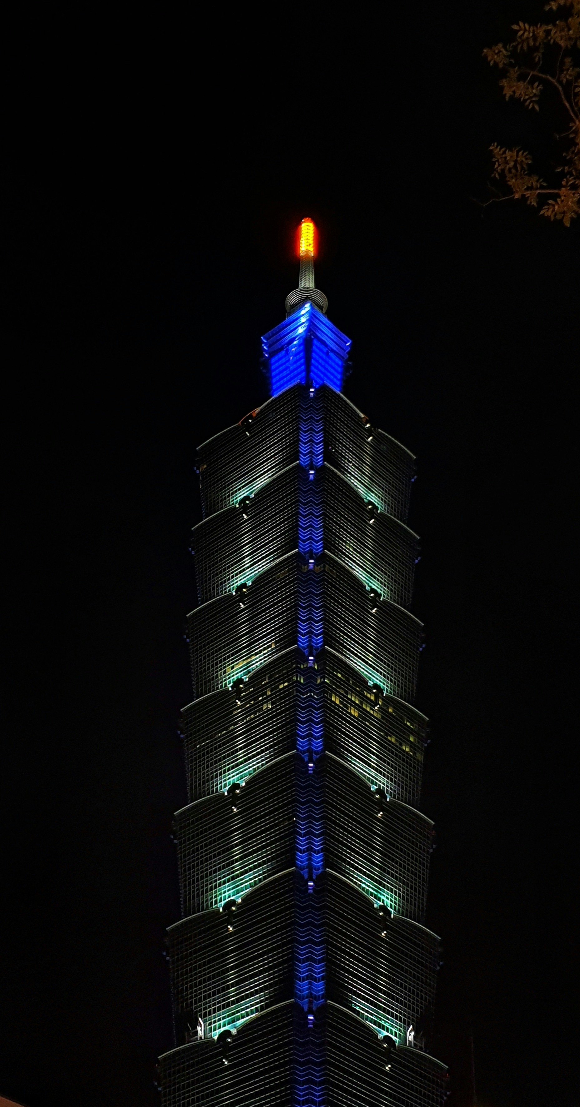Night photograph of Taipei 101 tower illuminated in blue with a red beacon at the tip. It showcases the tower's multi-tiered silhouette against a dark sky.