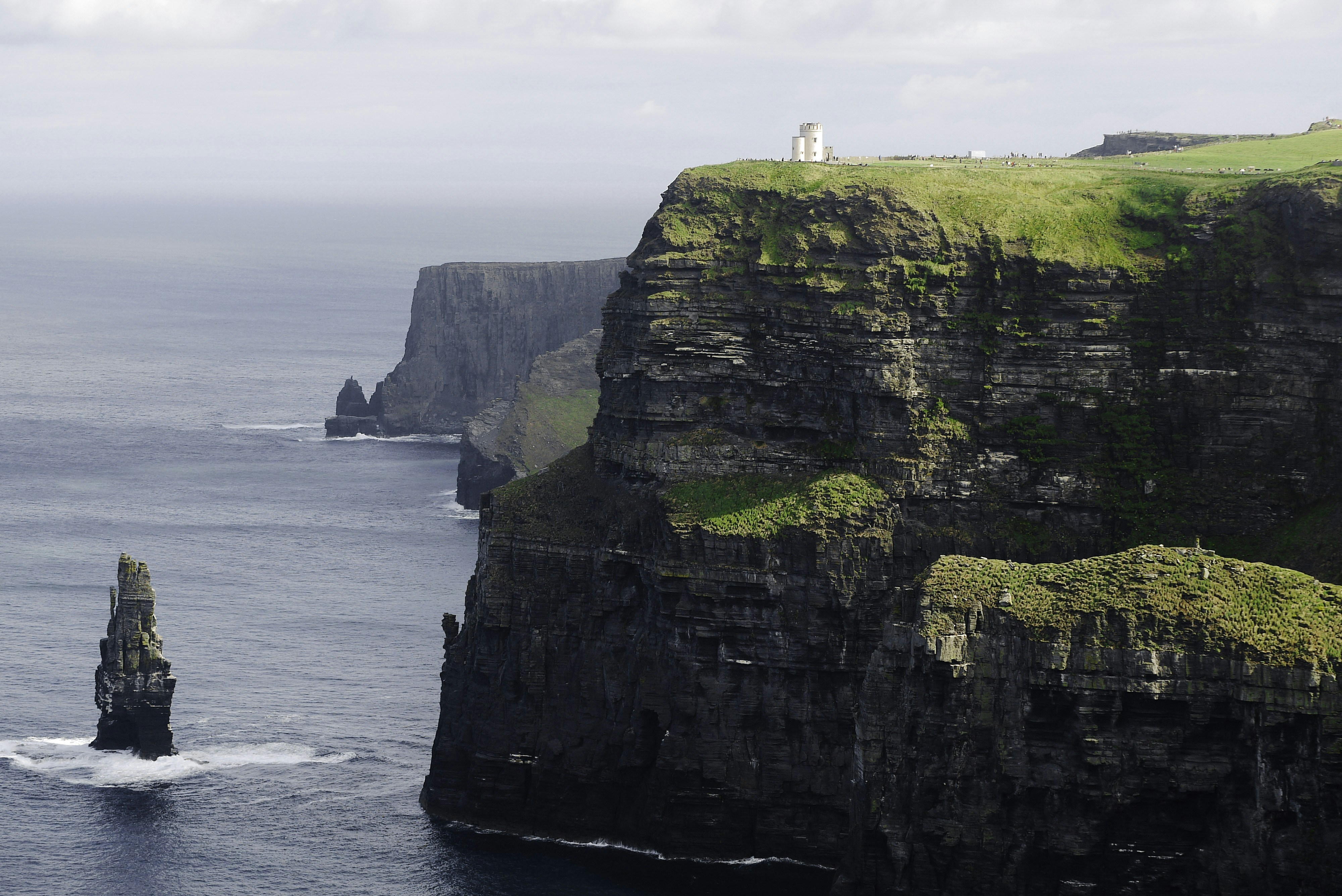 Dramatic cliffs rise from the ocean with a solitary tower perched atop, against a backdrop of serene waters and distant horizon.