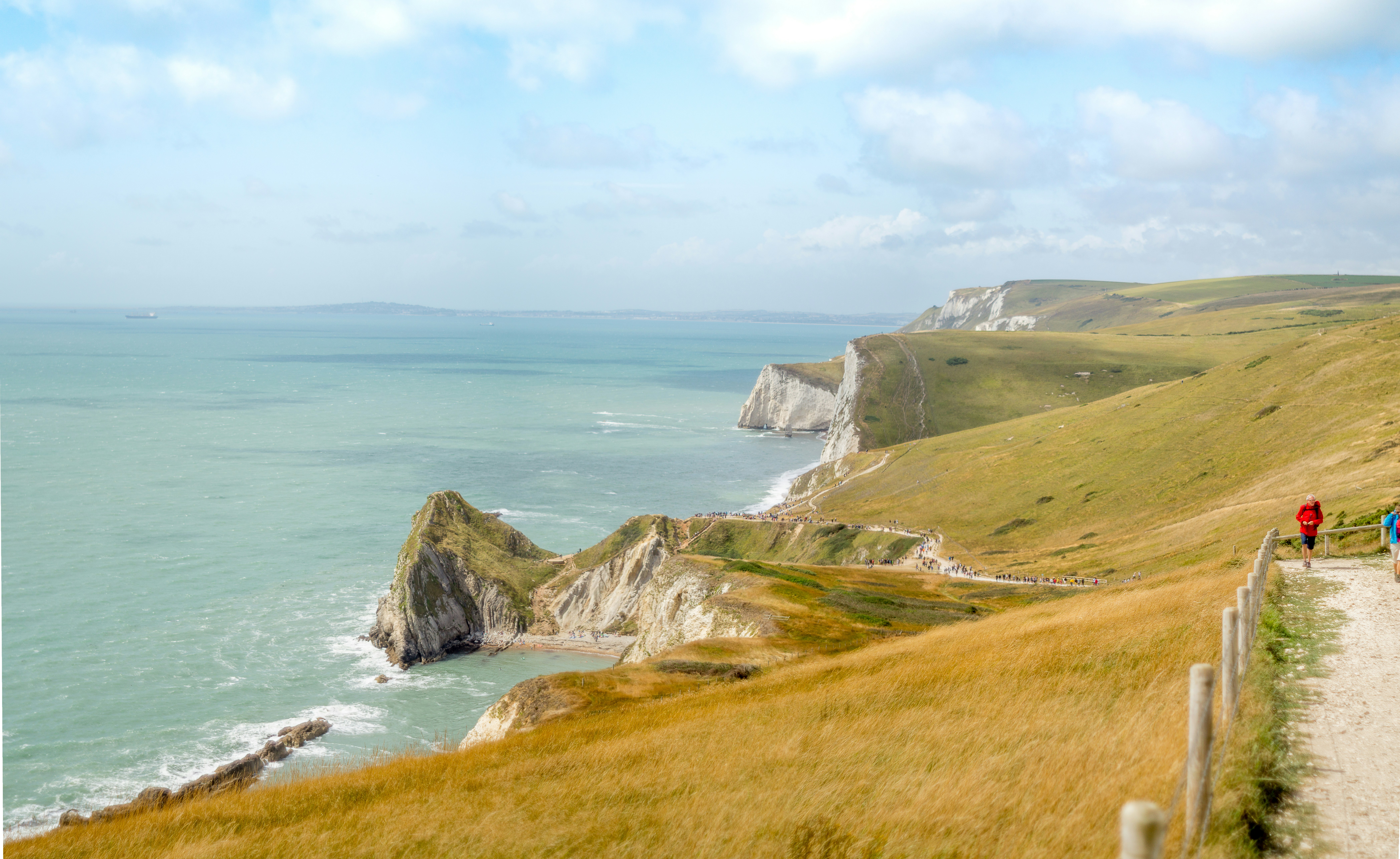 Coastal cliffs with a winding path overlooking a turquoise sea under a partly cloudy sky.