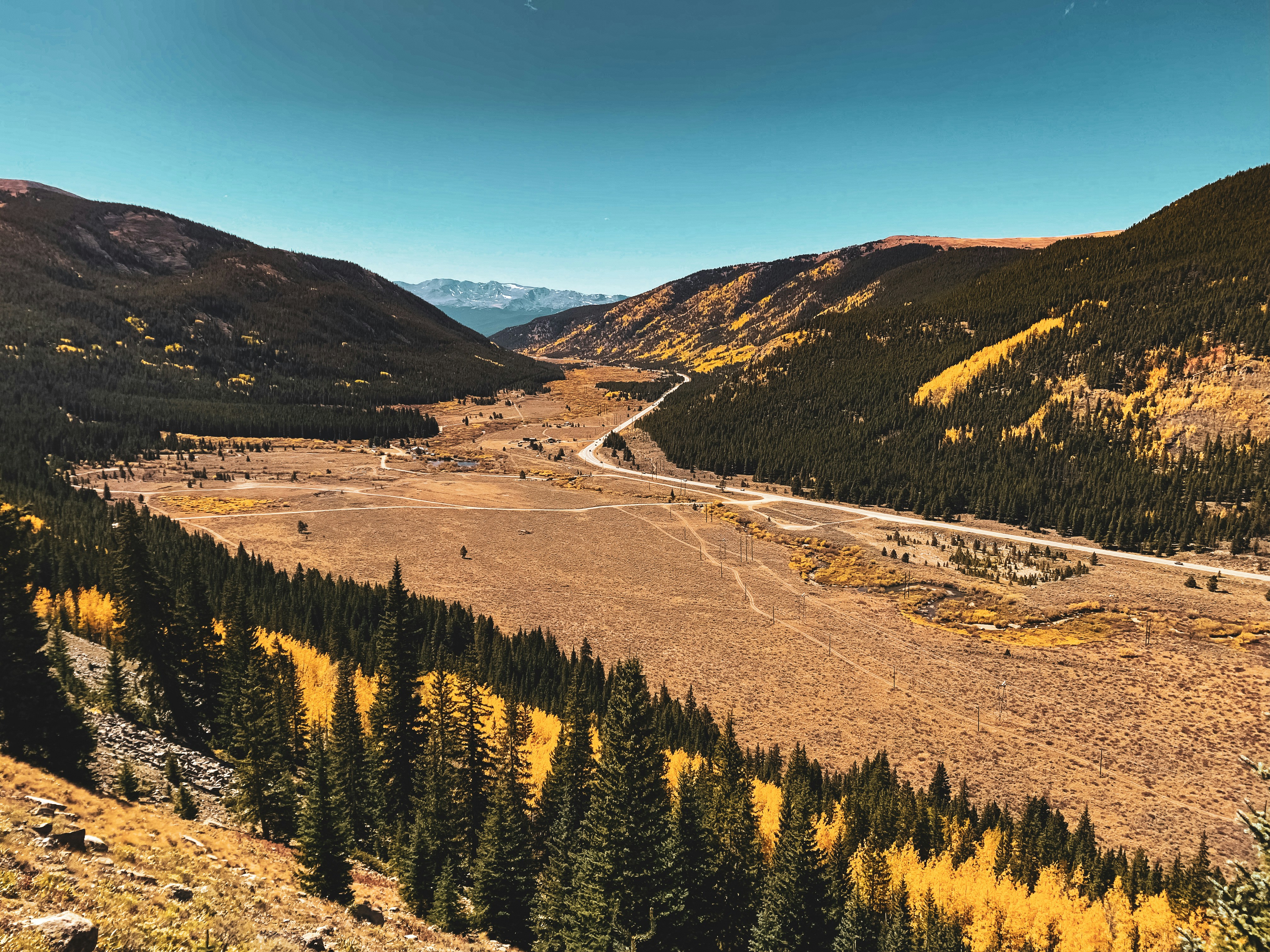 Vast valley surrounded by mountains, showcasing vibrant autumn foliage with golden aspens and lush evergreens. The winding road invites exploration.