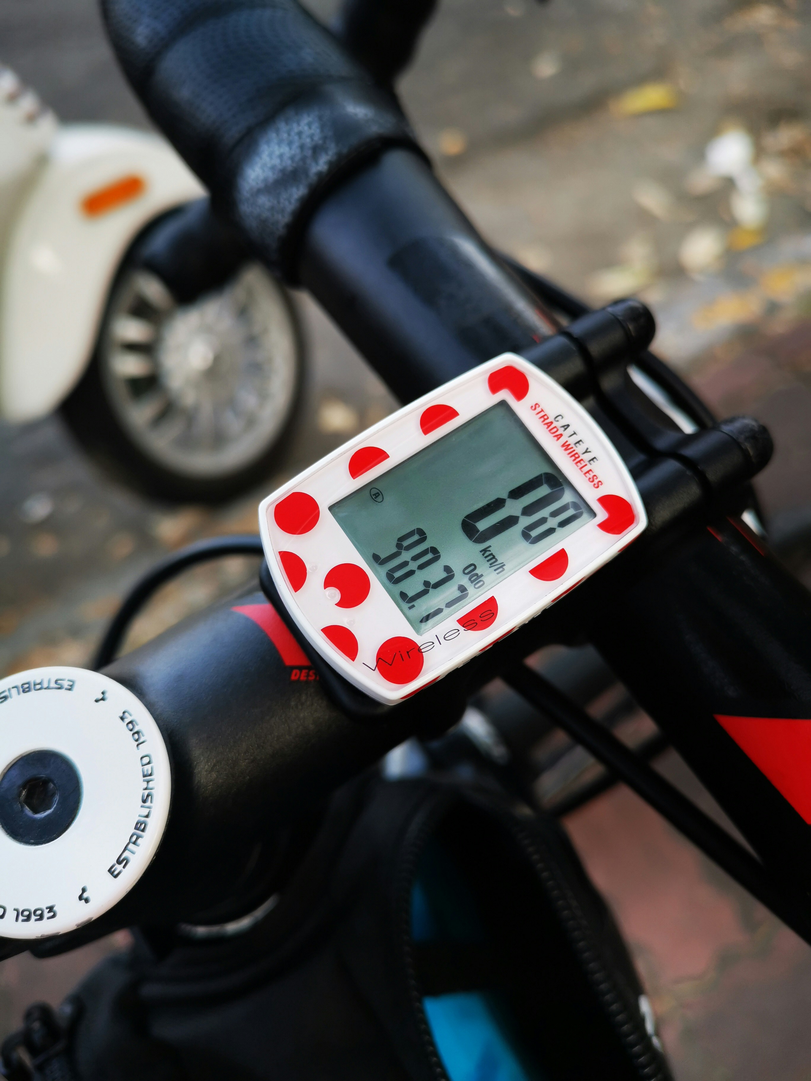 Close-up of a bike handlebar with a digital speedometer, handlebars and part of the frame in view; device shows numbers while urban environment blurs in the background.