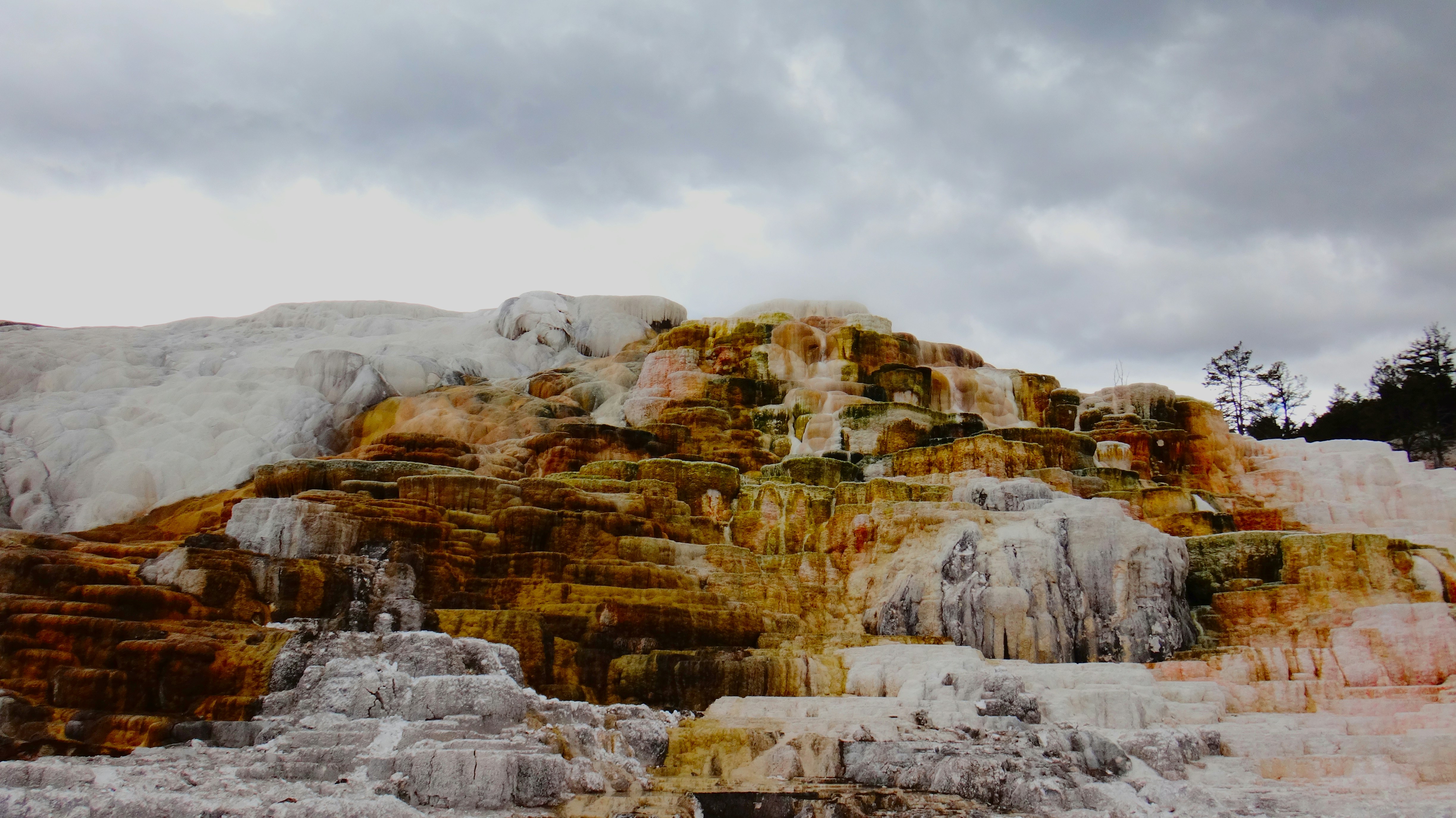 Vibrant mineral terraces display a spectrum of colors atop a rocky landscape, reflecting the natural beauty of geothermal activity. The cloudy sky adds a dramatic backdrop.