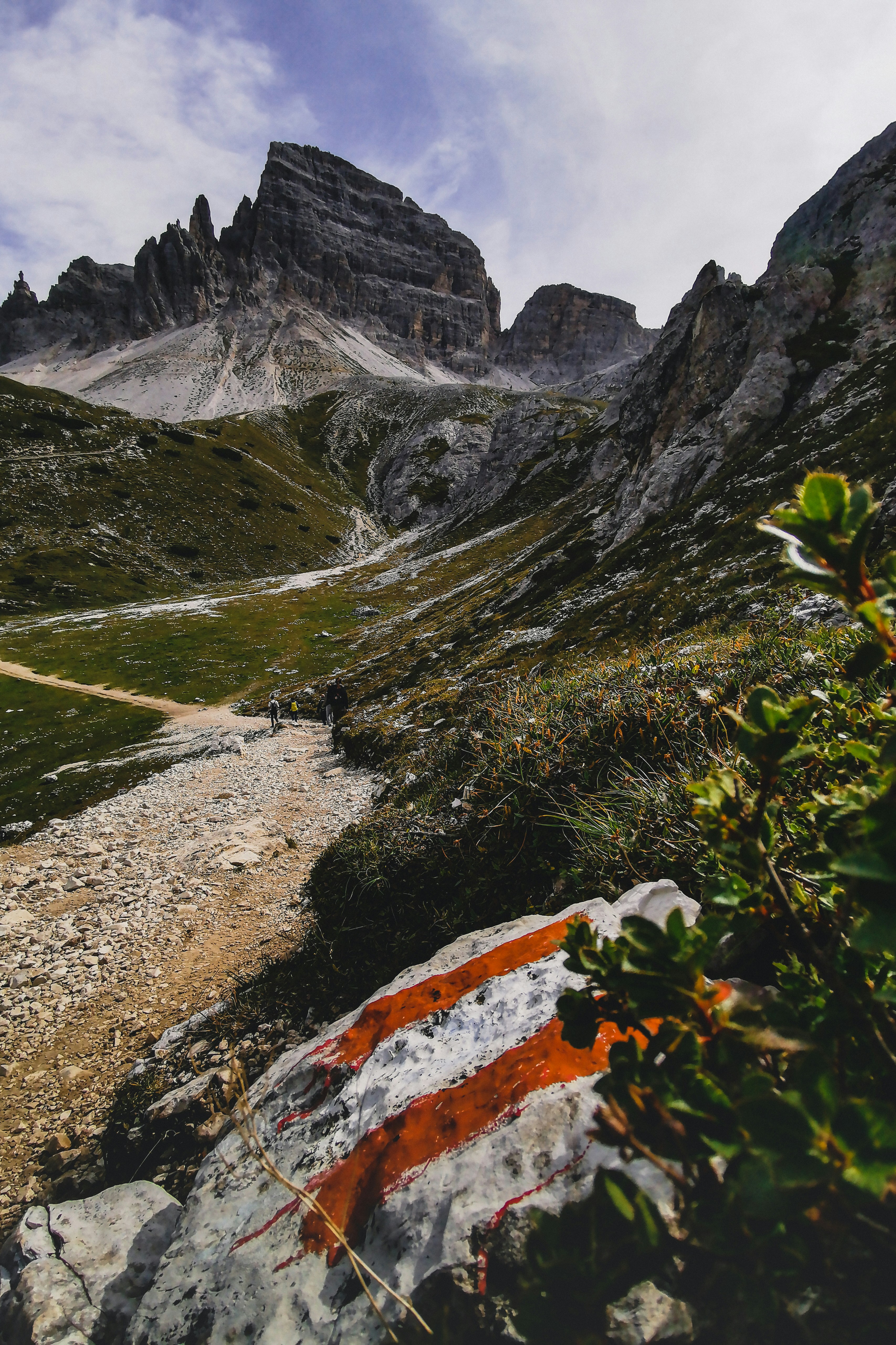 Rugged landscape photograph of a rocky trail winding toward jagged mountain spires under a partly cloudy sky, with hikers visible in the distance.