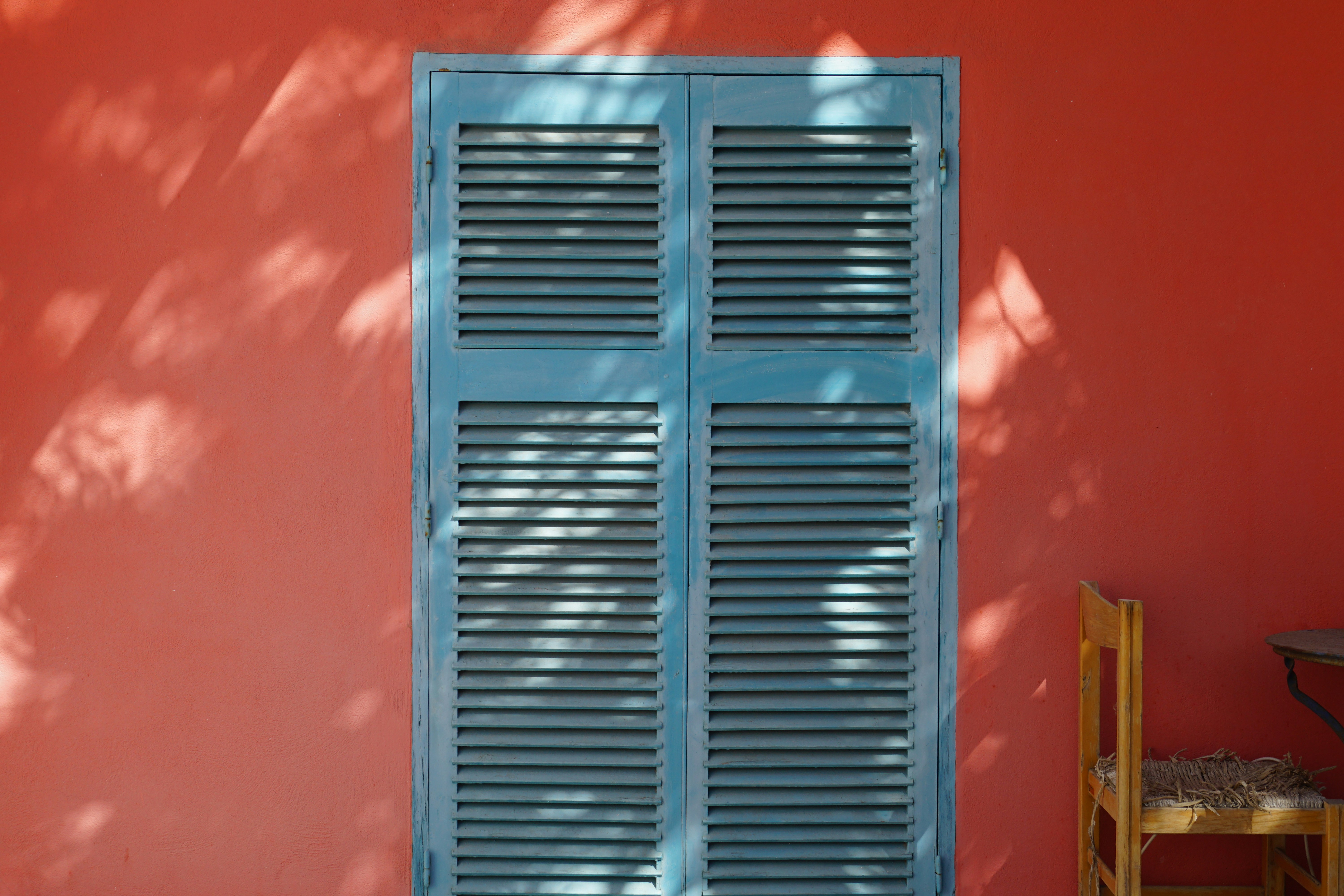 Blue wooden shutters against a vibrant red wall, casting intricate shadows, with a wooden chair nearby.
