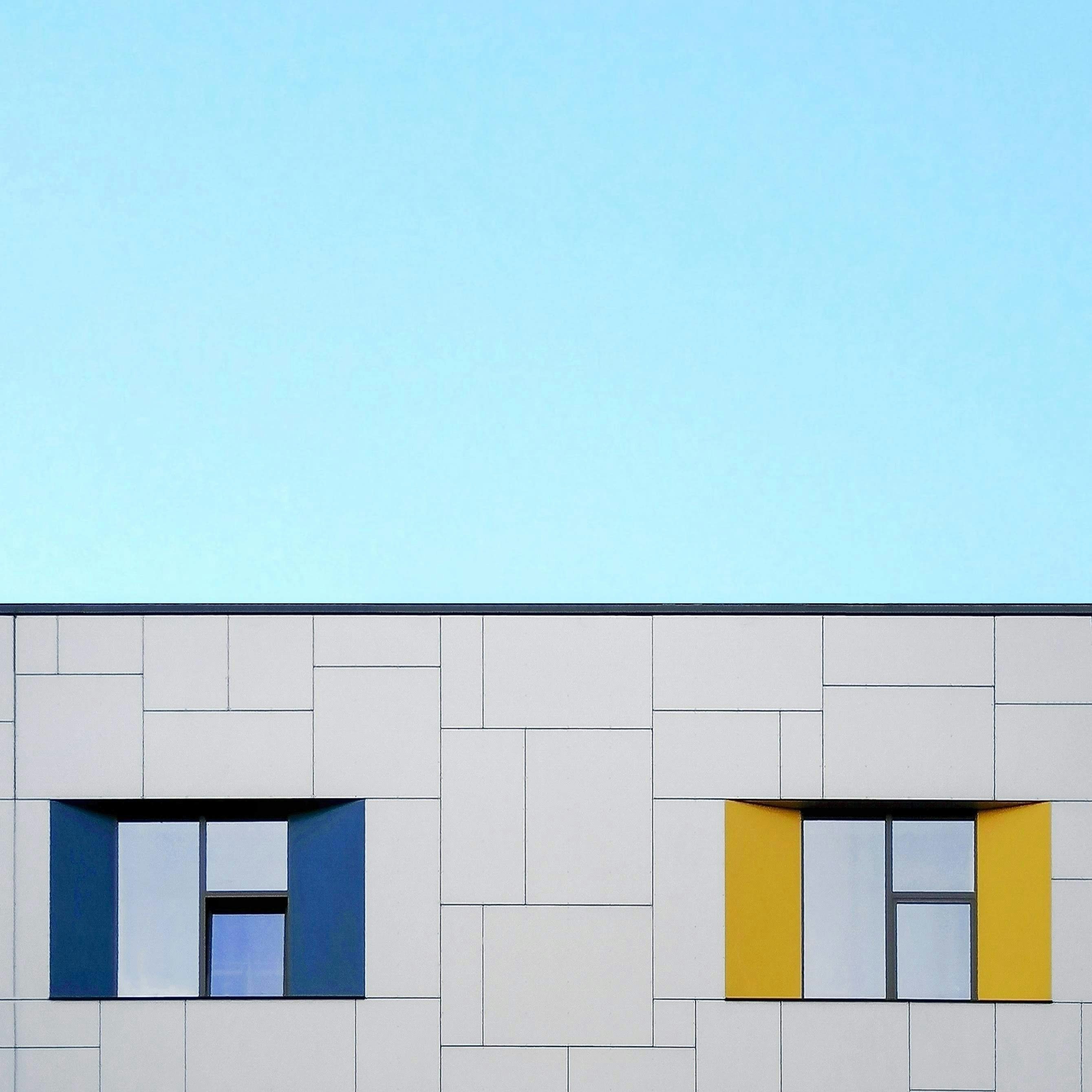 A modern building facade featuring blue and yellow window frames set in pale panels beneath a clear blue sky.