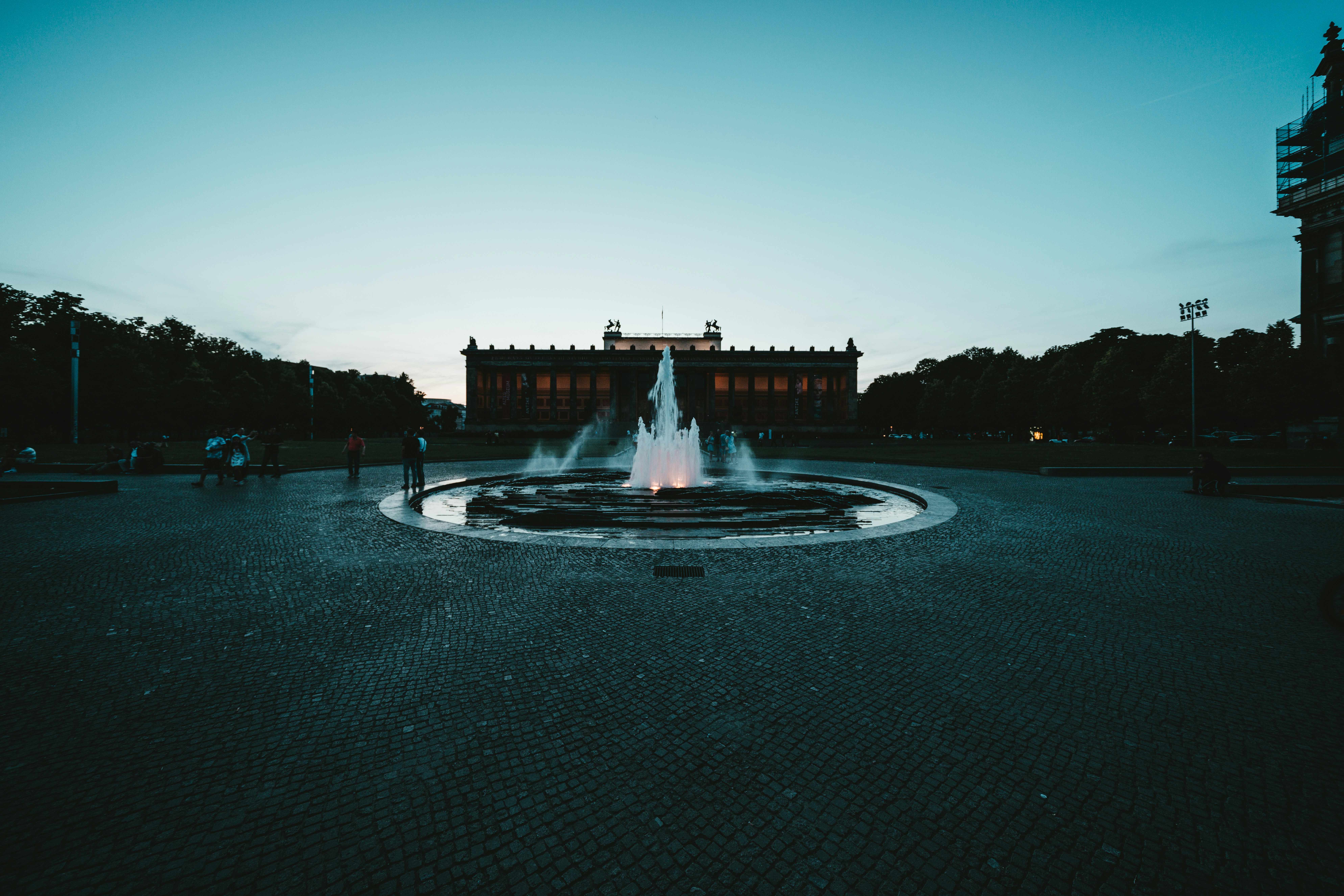 A tranquil fountain illuminated at dusk, surrounded by a spacious plaza and silhouetted by a historic building in the background.