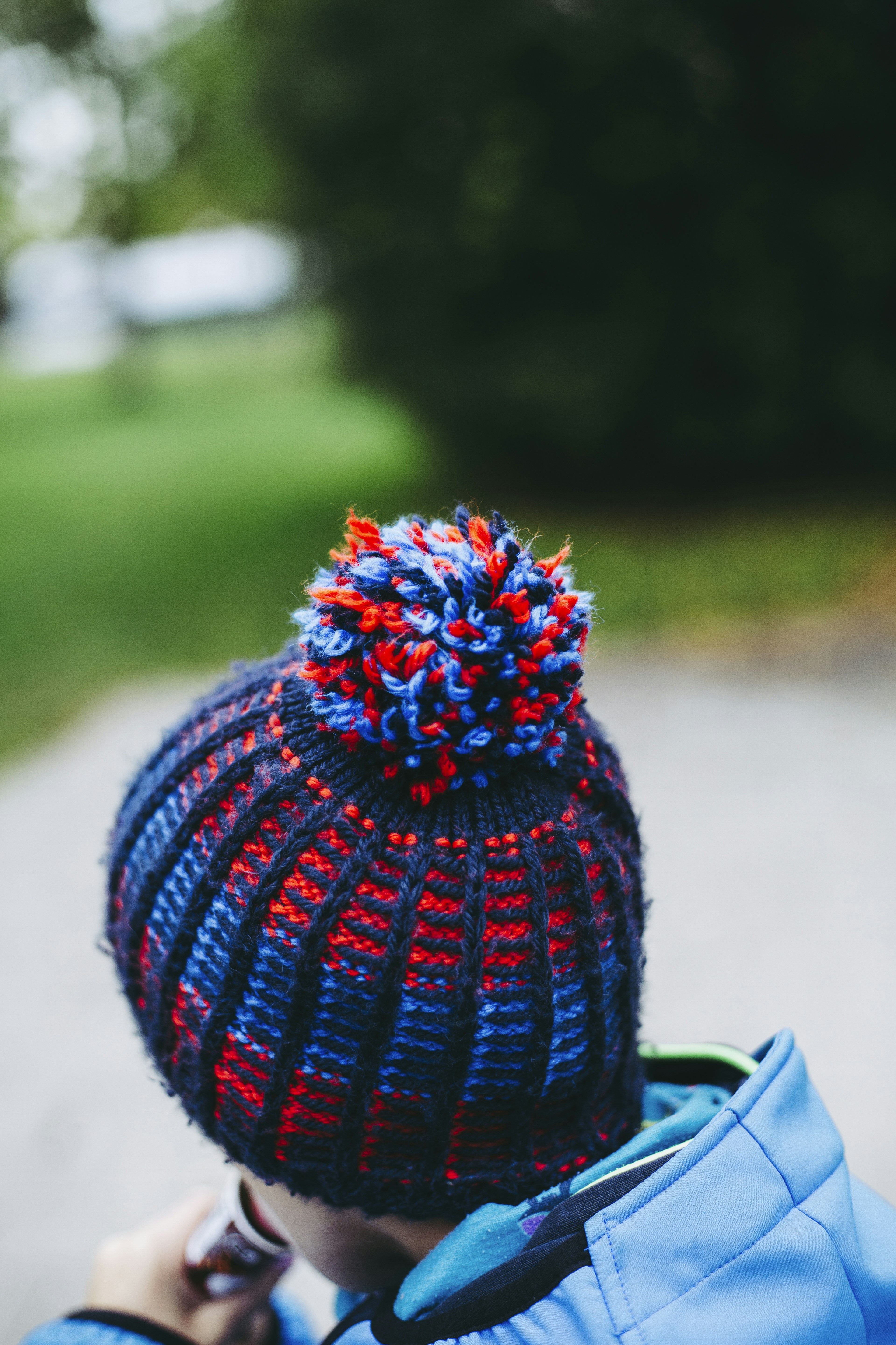 Child wearing a colorful knitted hat with a pom-pom, seated in a park setting. The vibrant colors contrast against the natural backdrop.