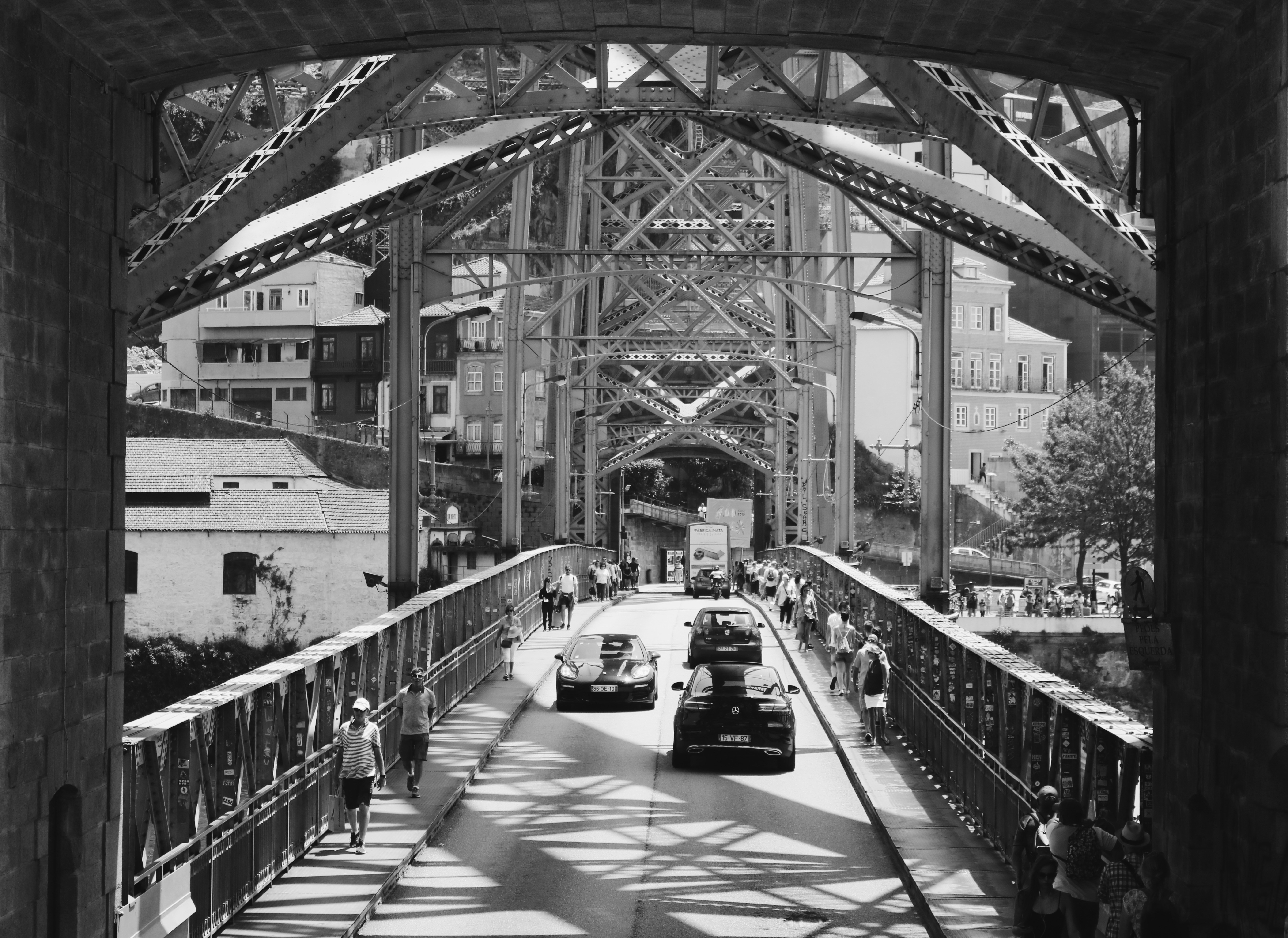 Black and white view of a bustling bridge with vehicles and pedestrians, framed by intricate steel architecture. The interplay of light and shadow creates a dynamic urban scene.