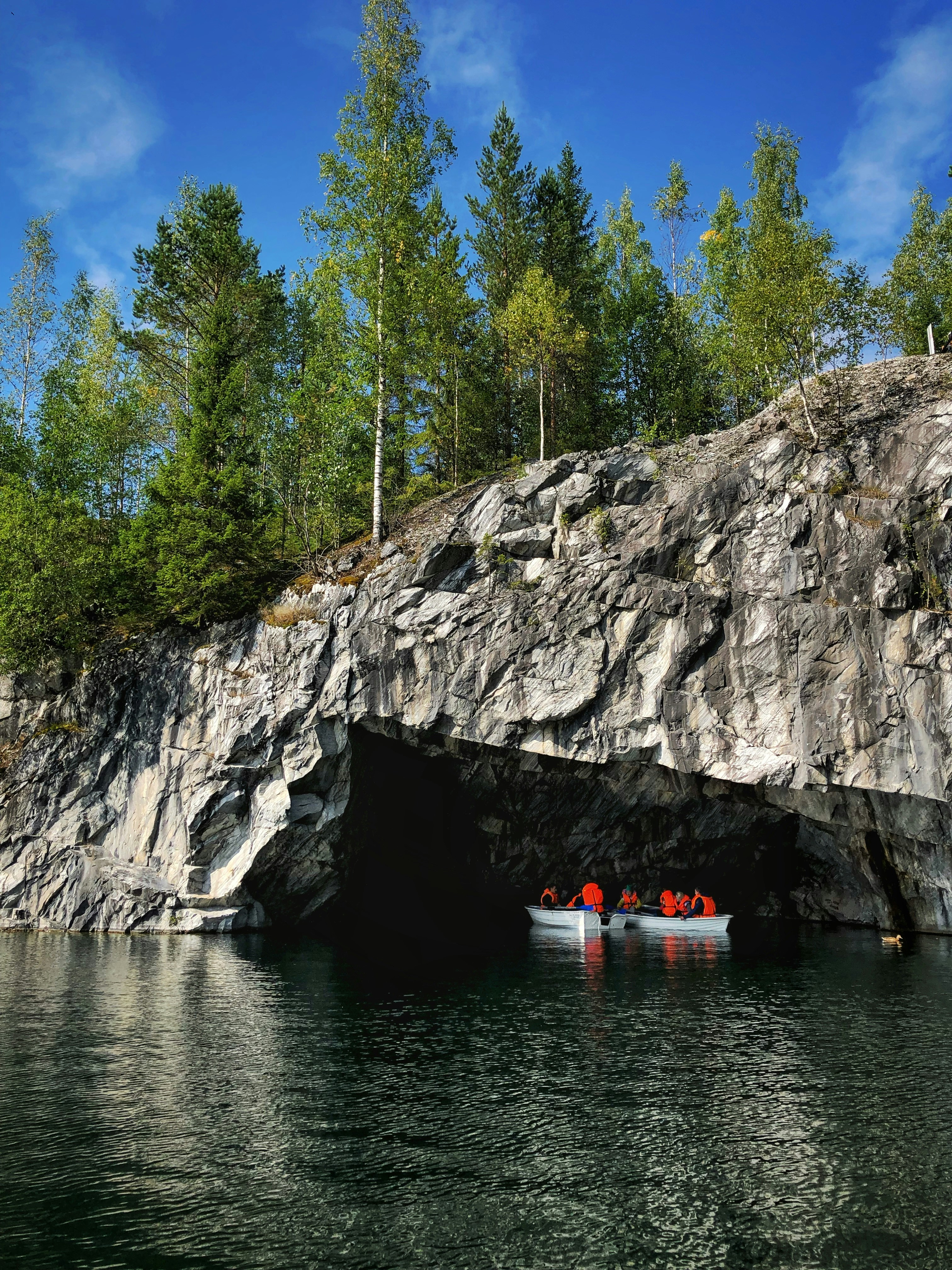 Kayakers in bright orange jackets navigate through a rocky cave by a serene lake, surrounded by lush greenery and clear blue skies.
