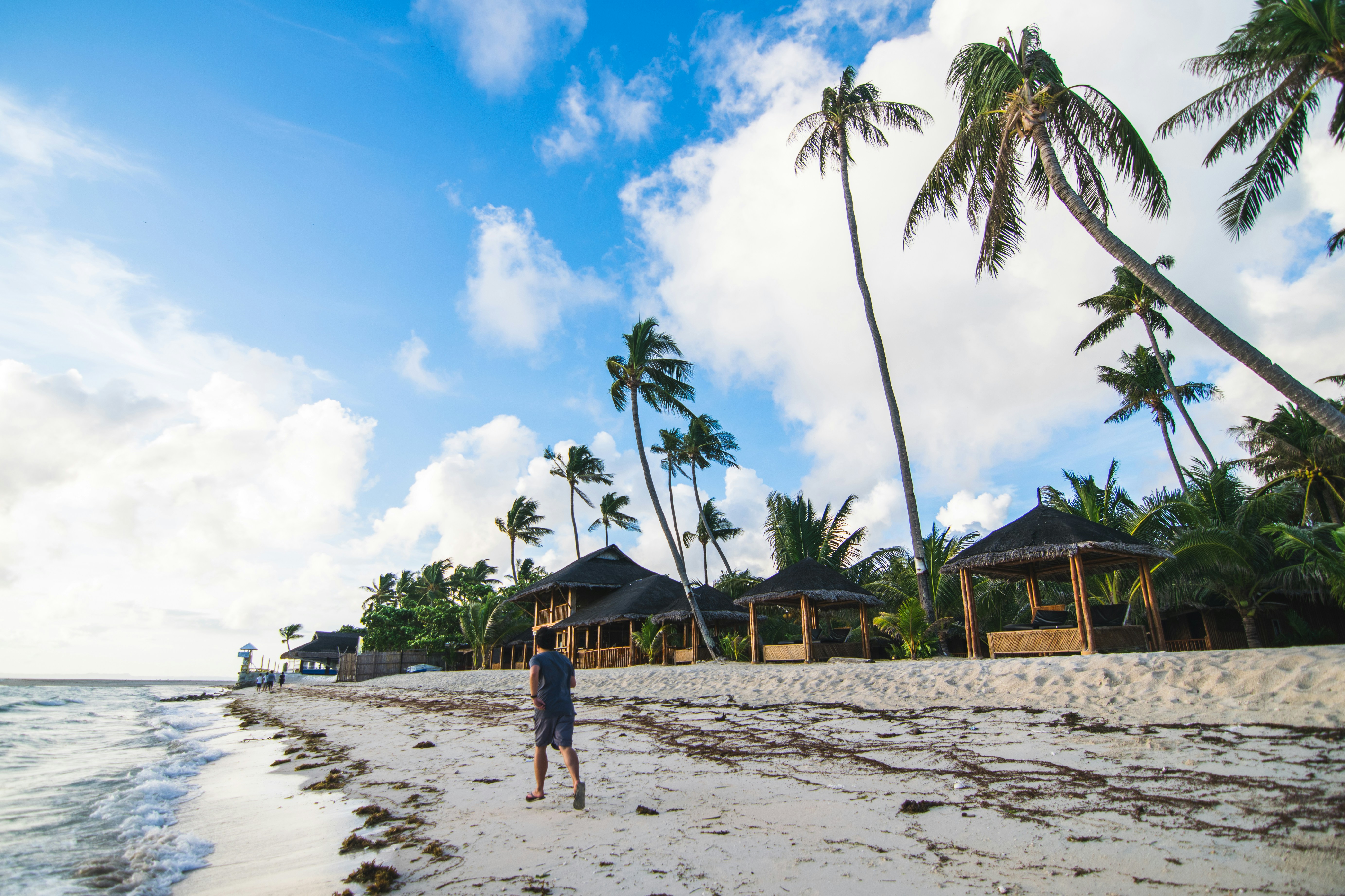 Person jogging on a sandy beach lined with palm trees and rustic huts under a bright blue sky.