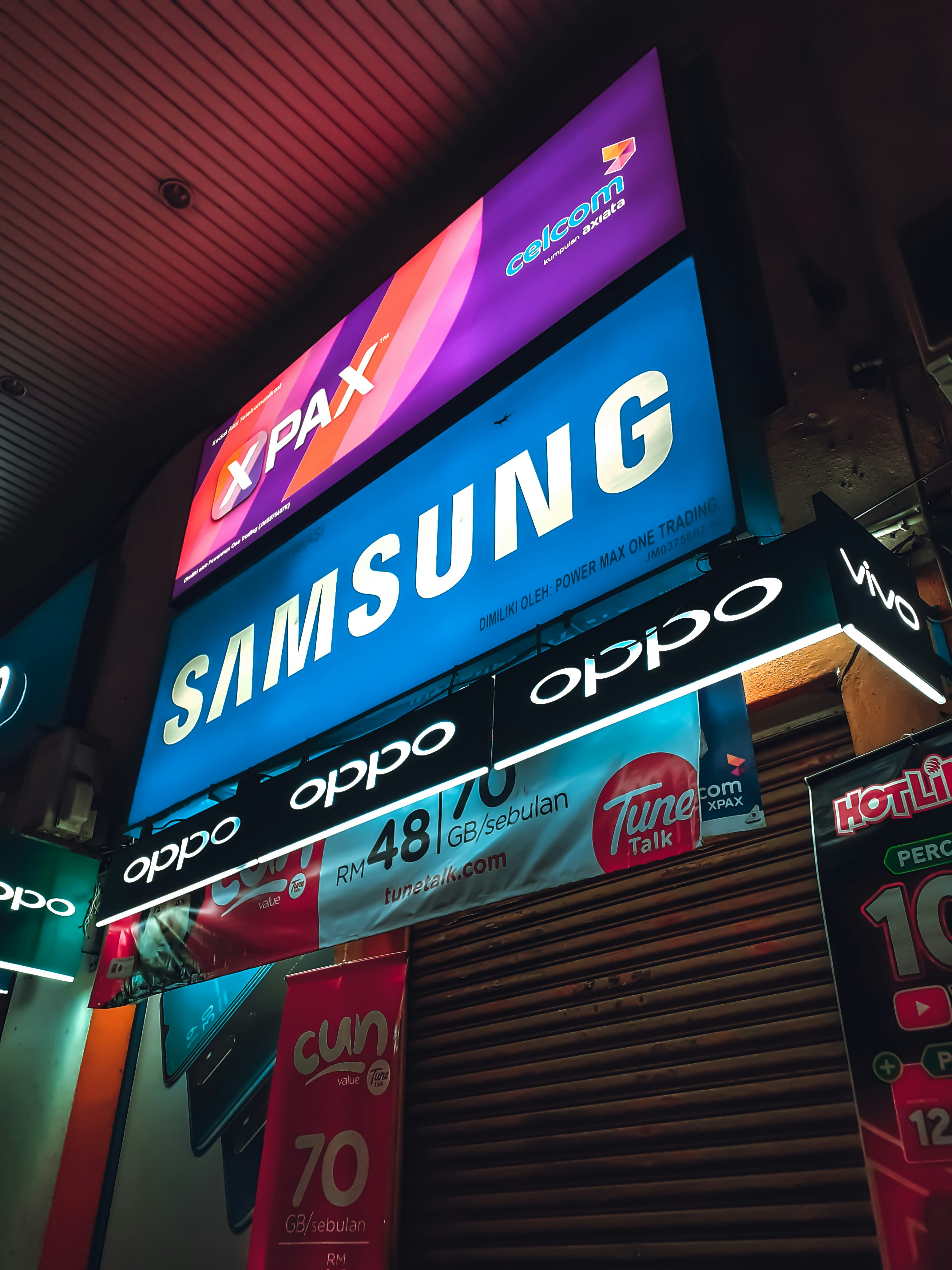 Vibrant advertising signs for Samsung and Oppo glow against a dark urban backdrop, highlighting the competitive mobile market. The scene captures the essence of city life at night.