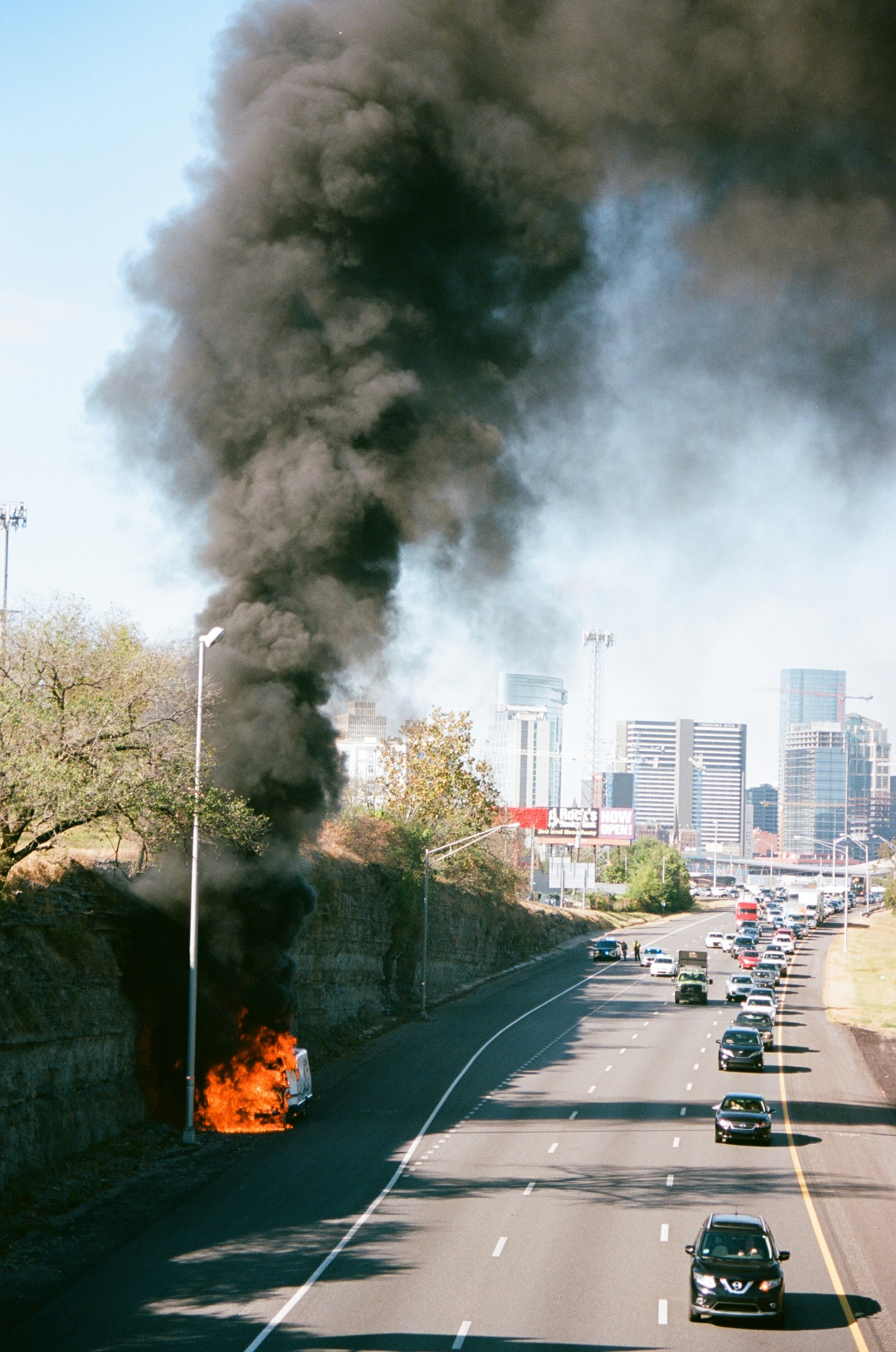 Dark plume from a fire along a multi-lane highway with a line of cars and cityscape in the distance.