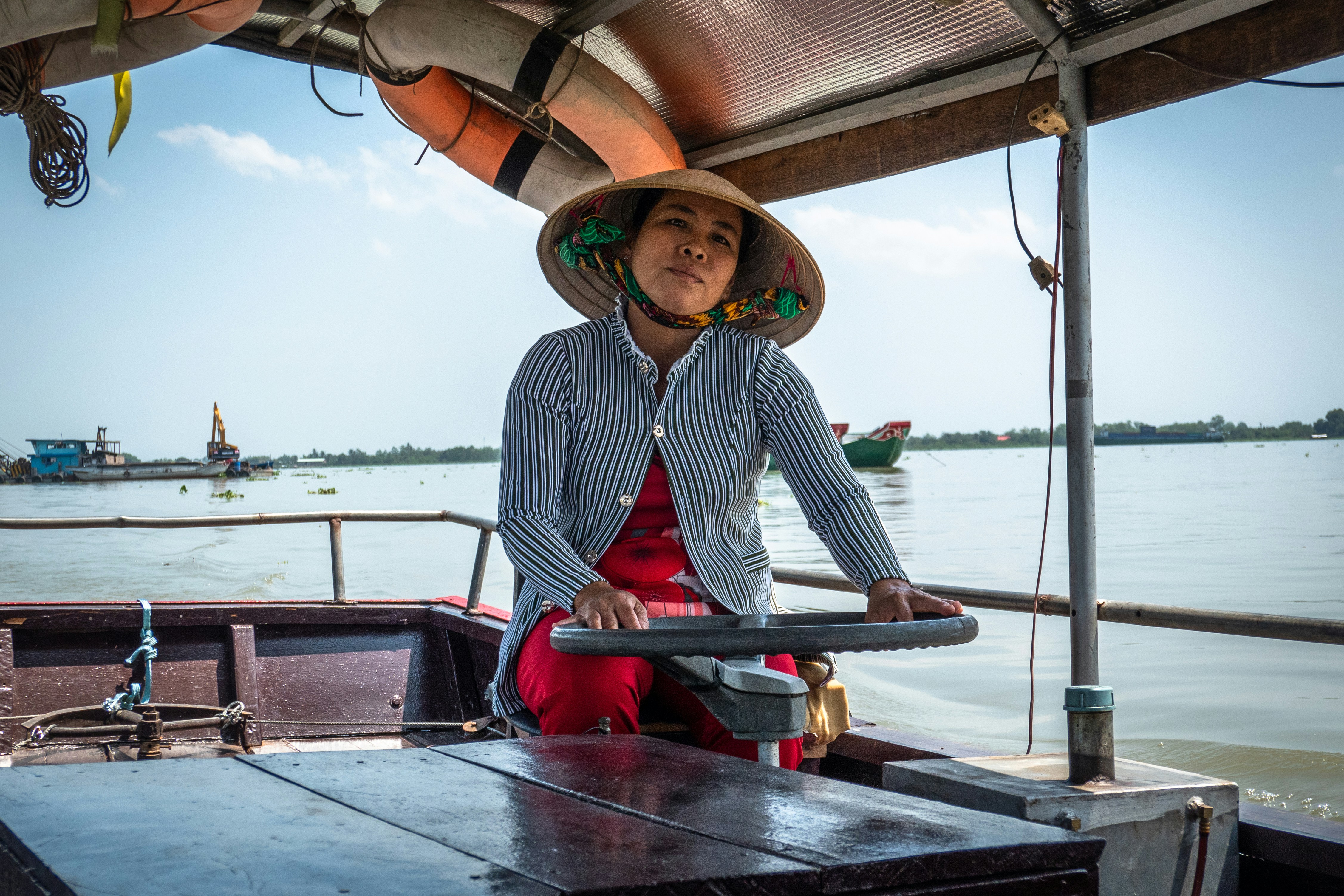 Woman steering a boat on the Mekong River under a clear blue sky.