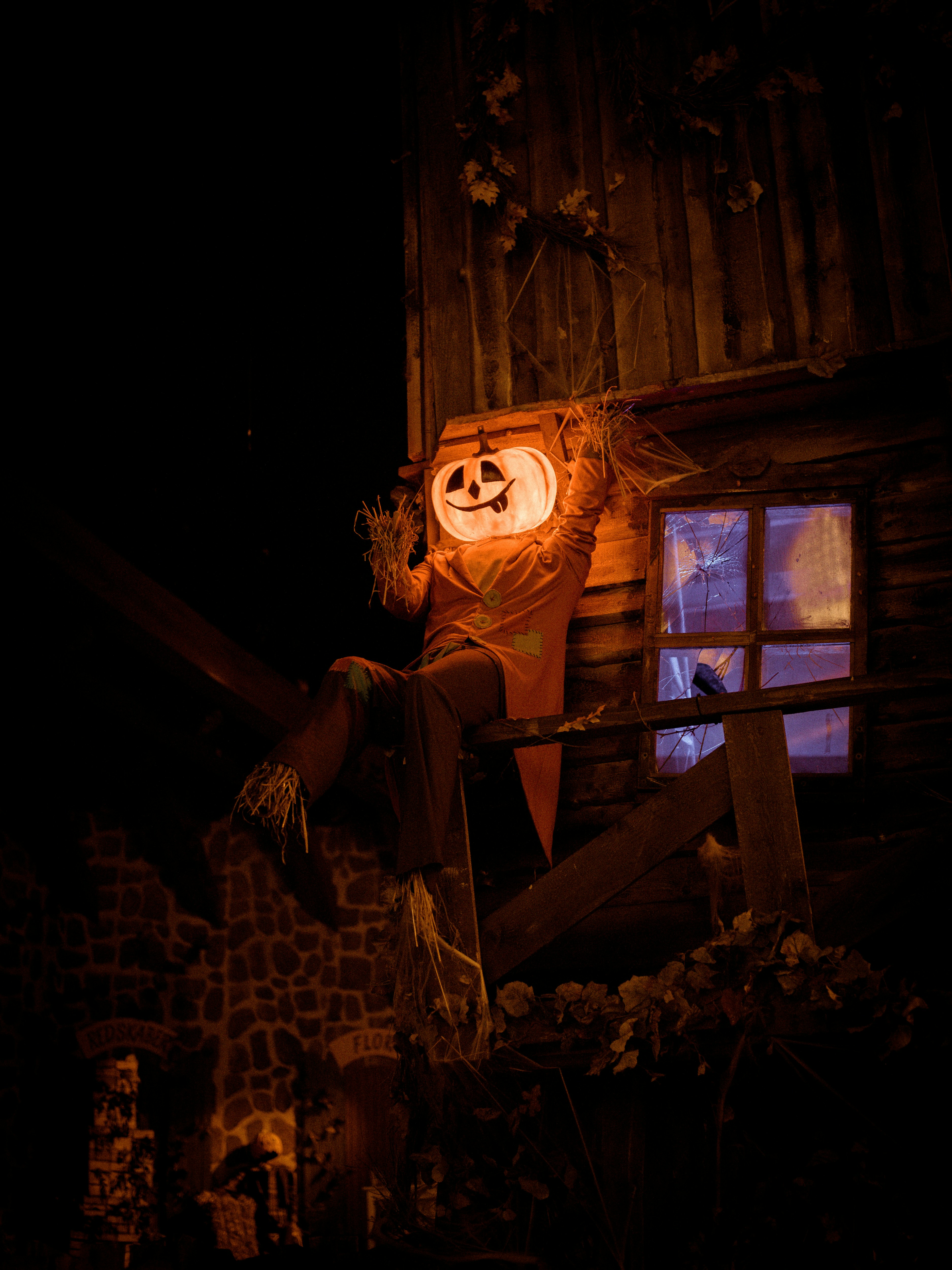 A whimsical scarecrow with a pumpkin head sits perched on a wooden railing, overlooking a dimly lit barn scene adorned with autumn decor.