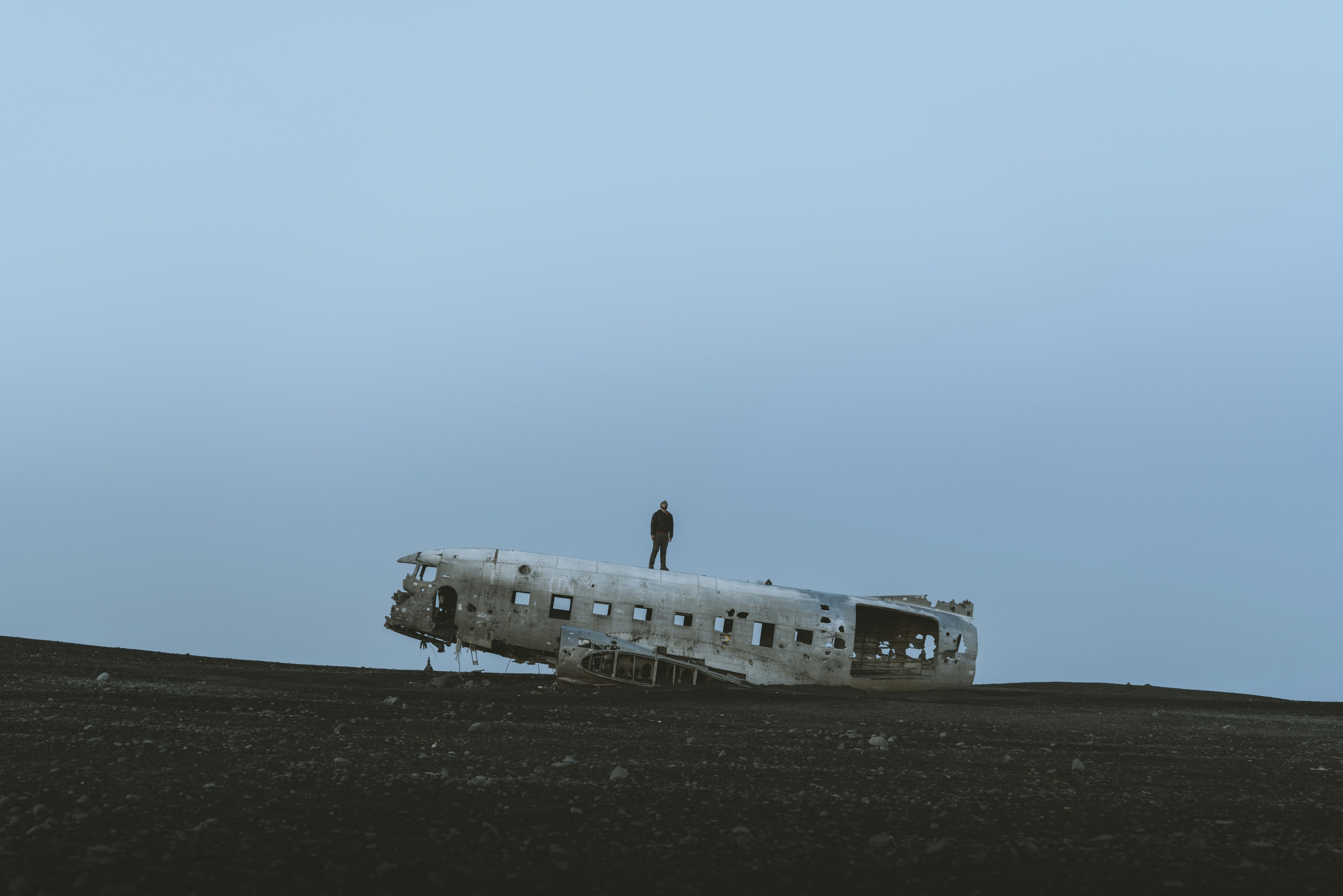 A lone figure stands atop a weathered airplane wreck on a desolate landscape, evoking themes of solitude and abandonment.