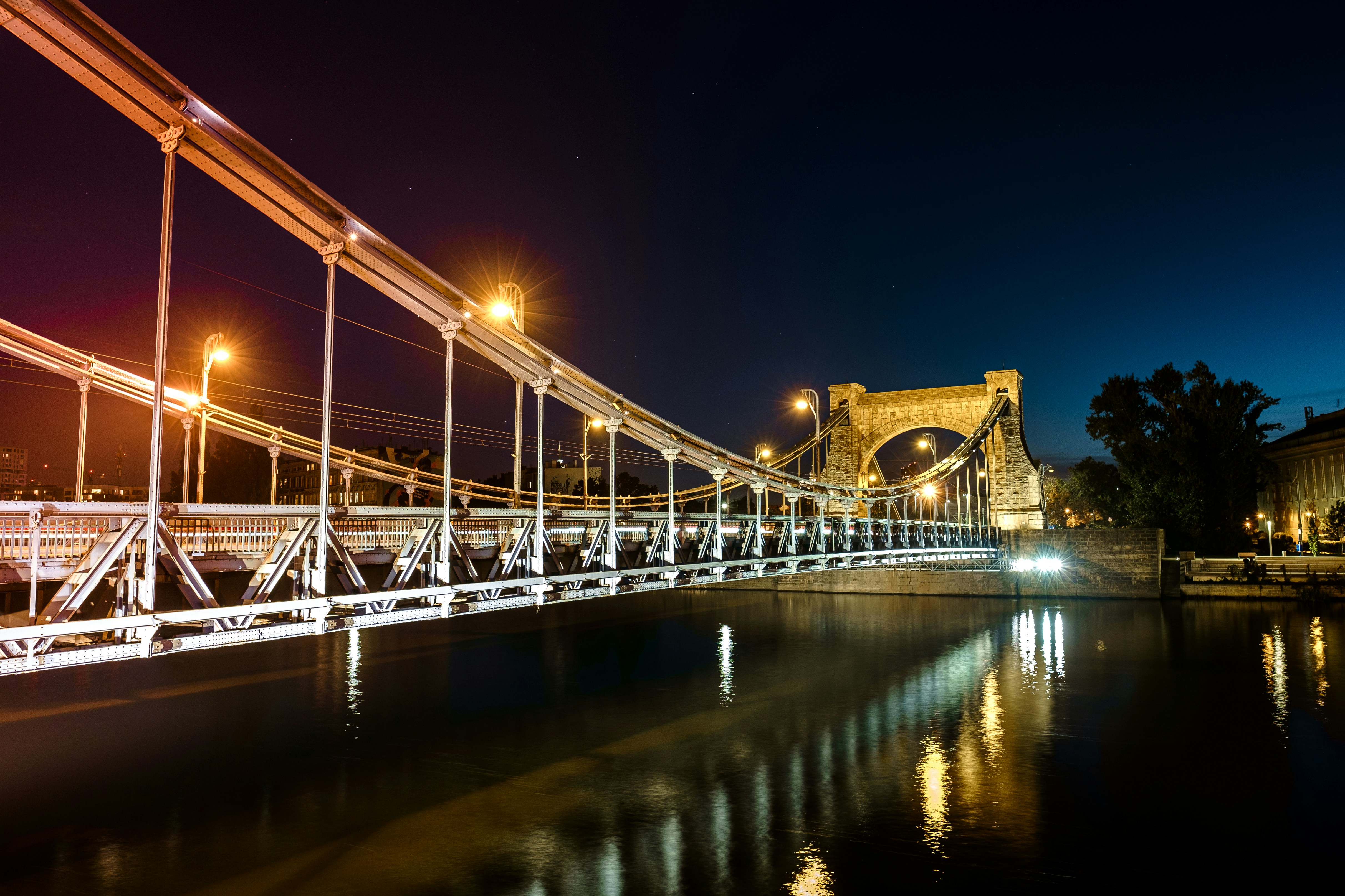 Illuminated bridge reflecting in calm waters under a night sky.