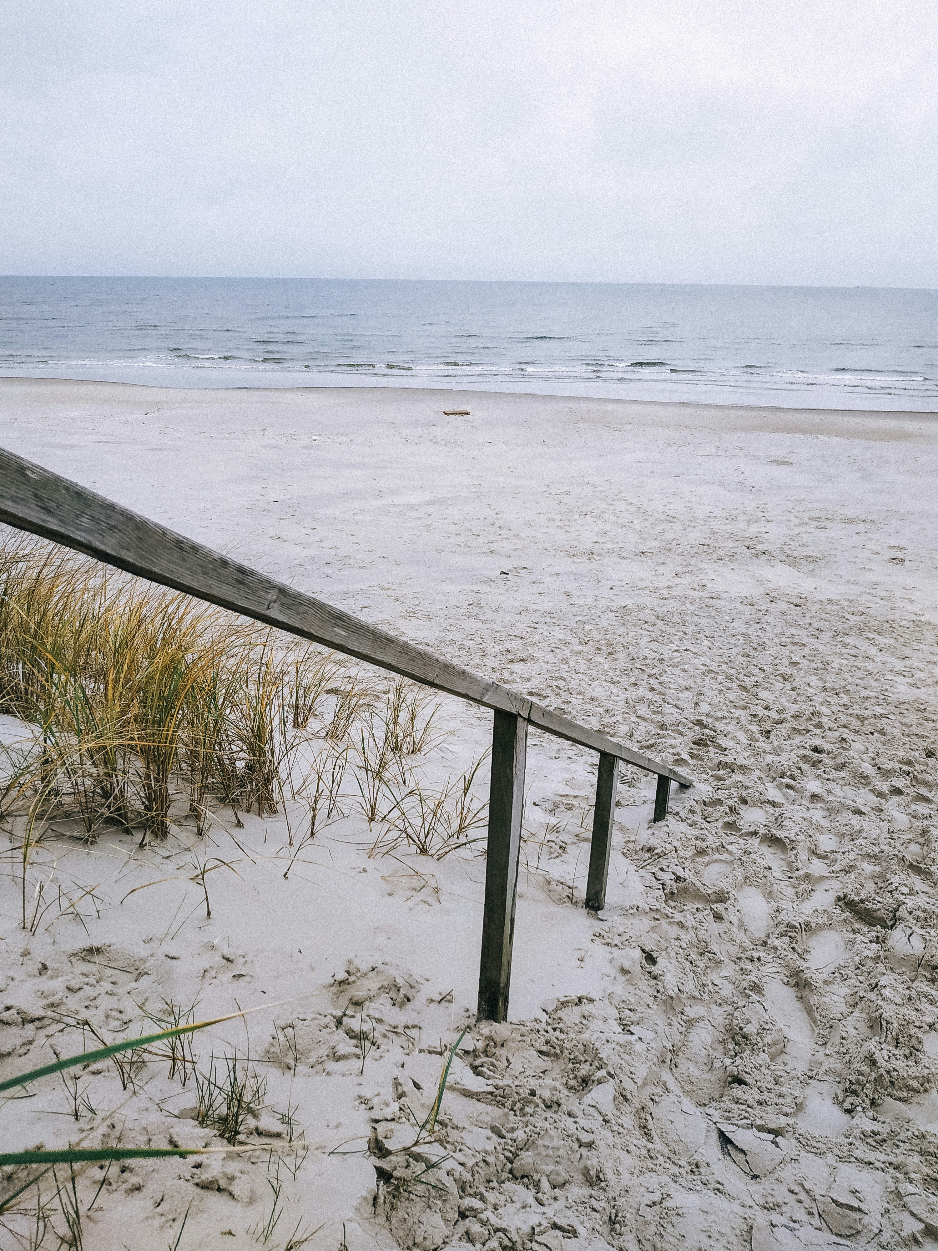Wooden ramp leading down to a sandy beach with gentle waves lapping at the shore, framed by beach grass. A tranquil coastal scene.