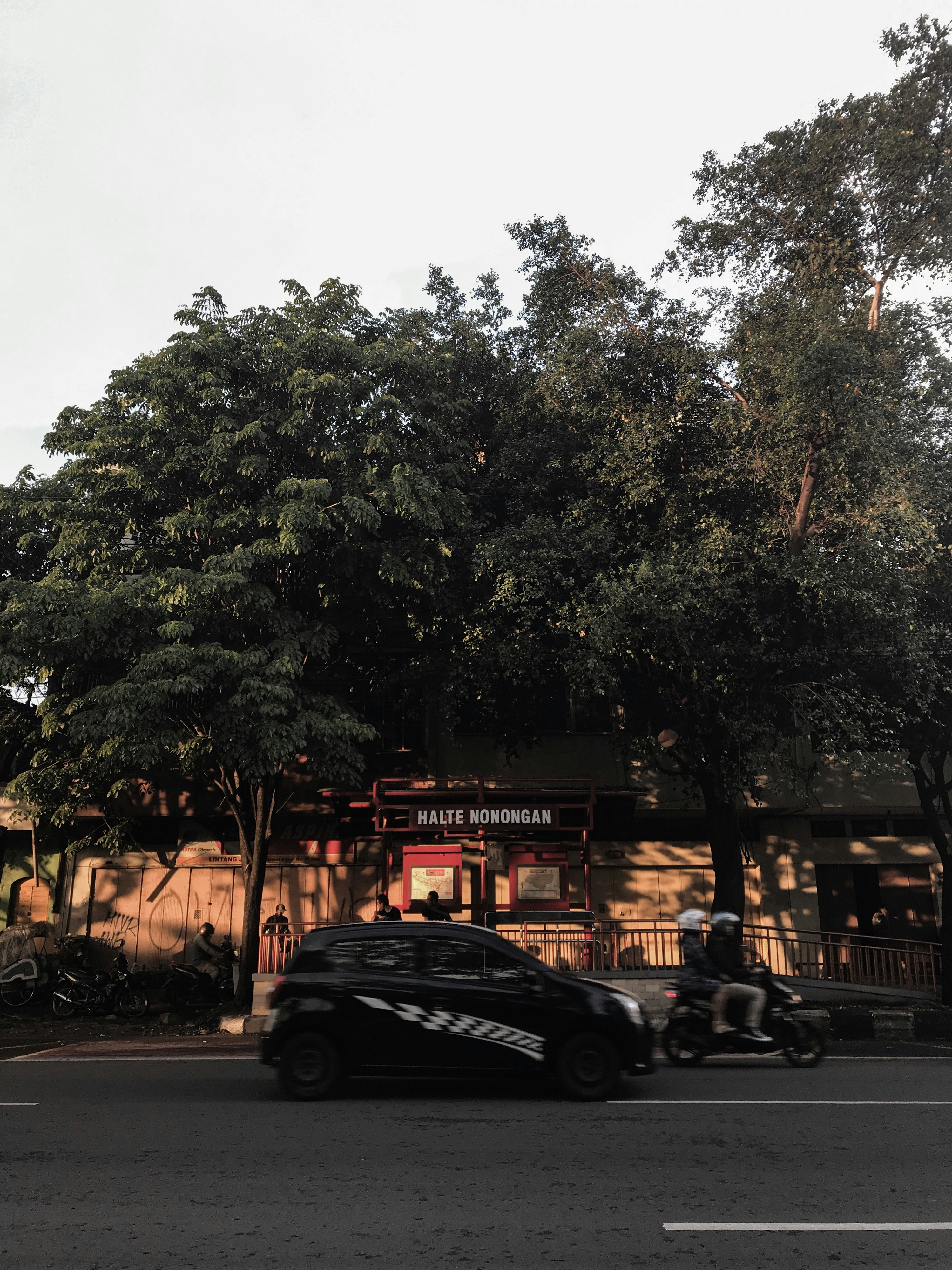 A bus stop named 'Halte Nonongan' nestled among lush trees, with a moving vehicle in the foreground. The warm light casts a serene ambiance.