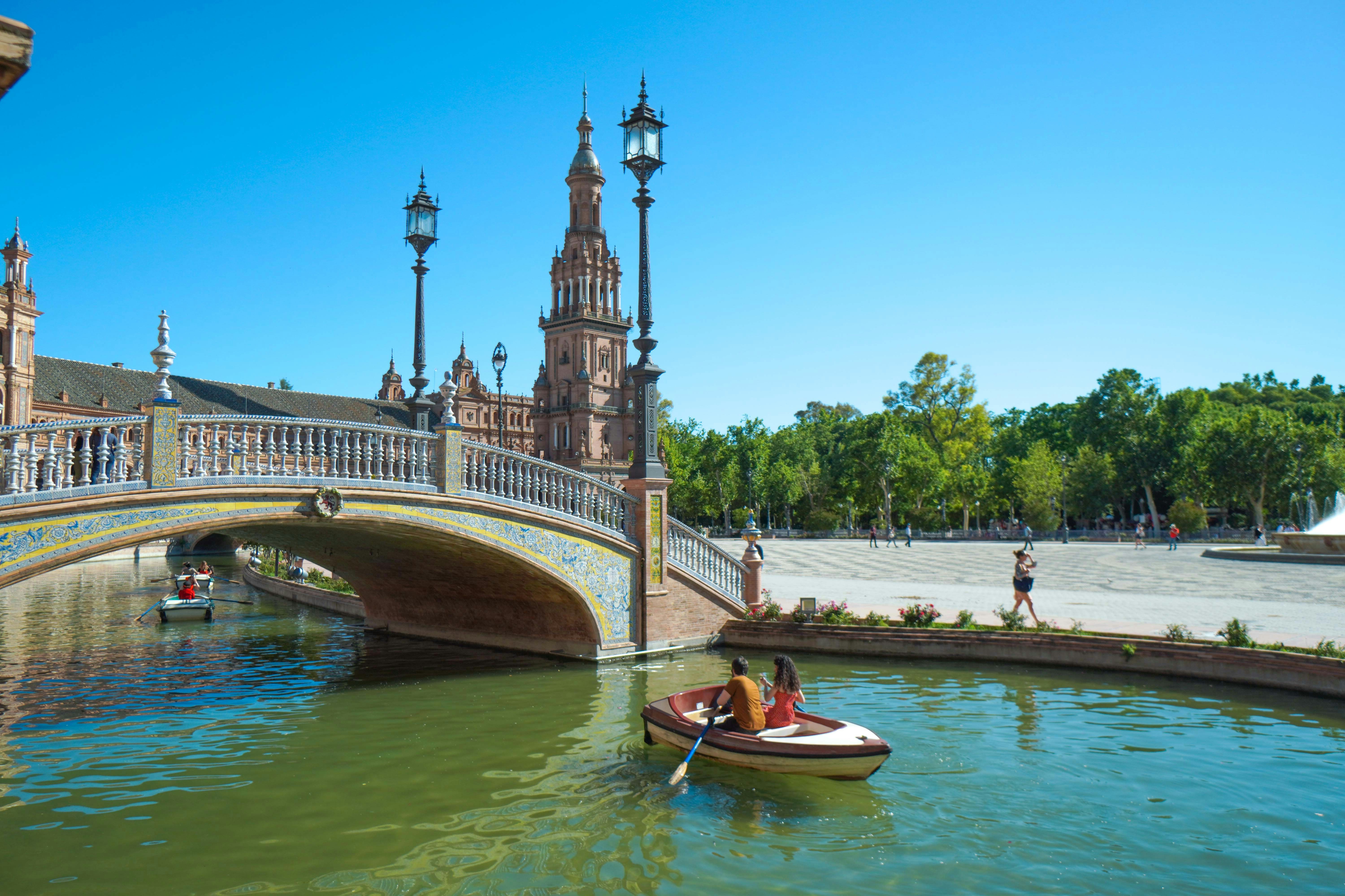 Couple rowing a boat beneath a decorative bridge in Plaza de España with historic towers and clear blue sky.