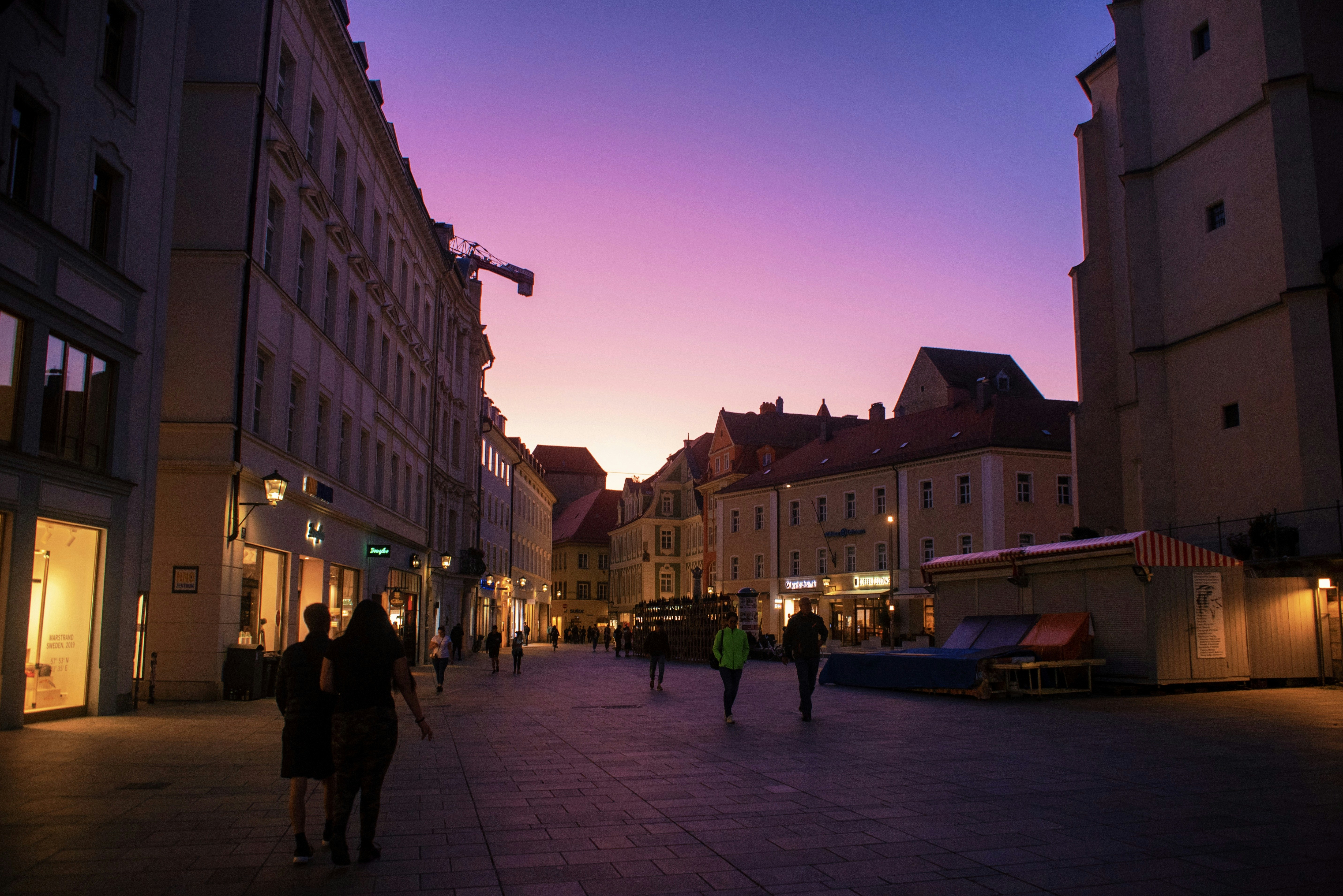 Evening scene in a charming European square, featuring people walking amidst beautifully lit buildings under a gradient sky. 