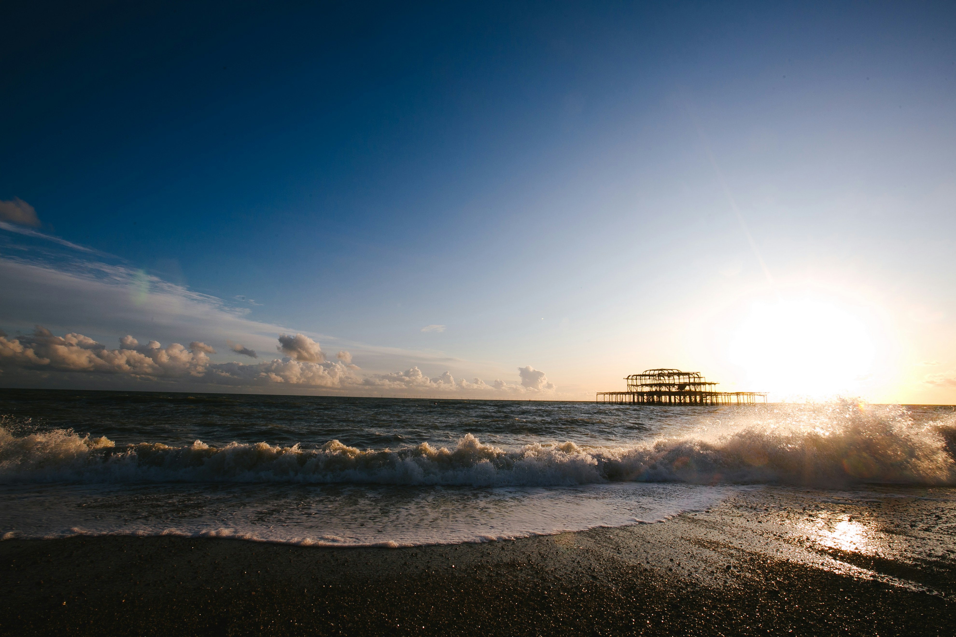 Ocean waves break against the shore under a vibrant sunset sky, with silhouetted structures in the distance.
