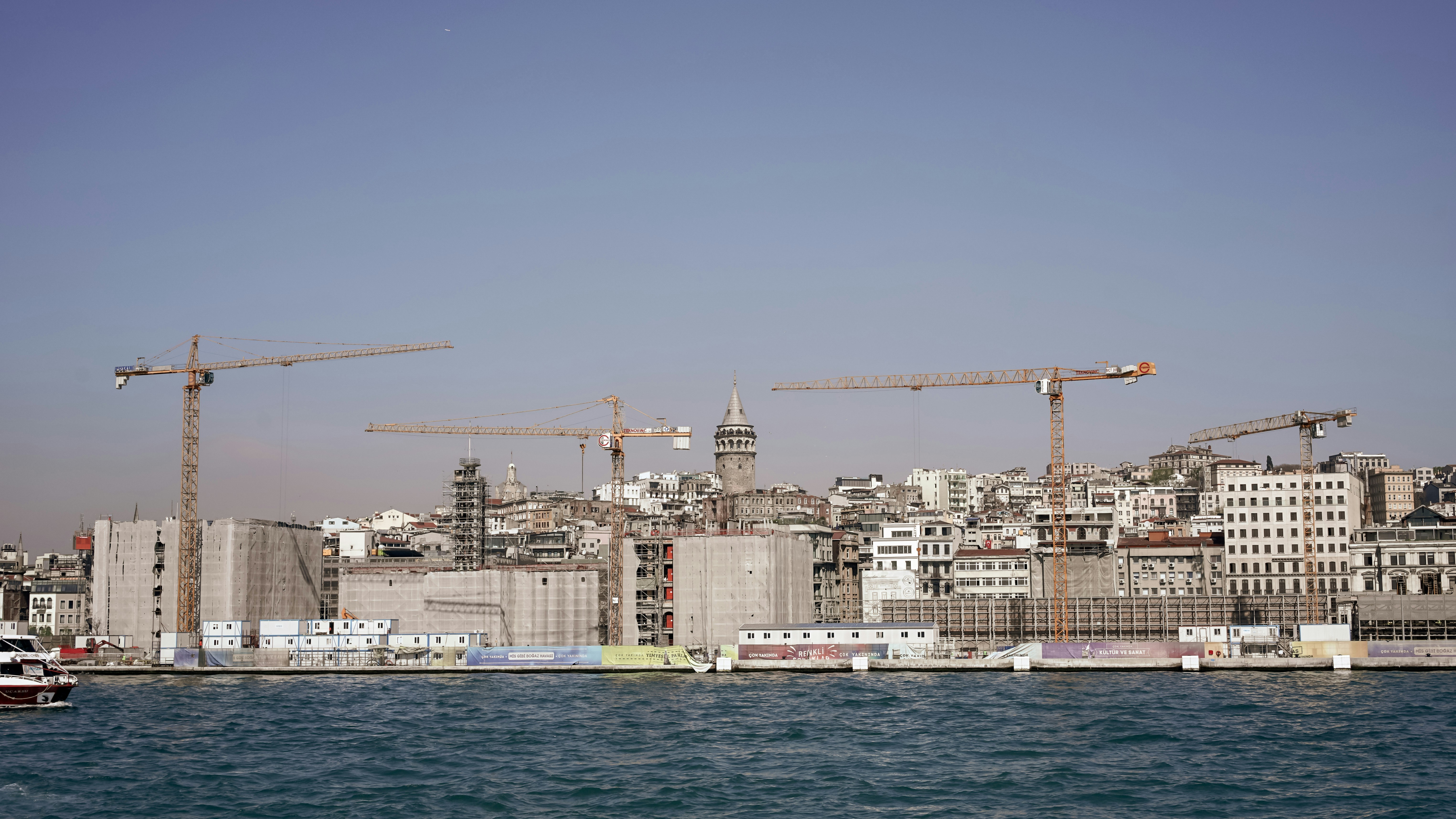 Lifelike city skyline along the harbor with multiple cranes towering over mid-rise buildings and a calm waterline in the foreground.