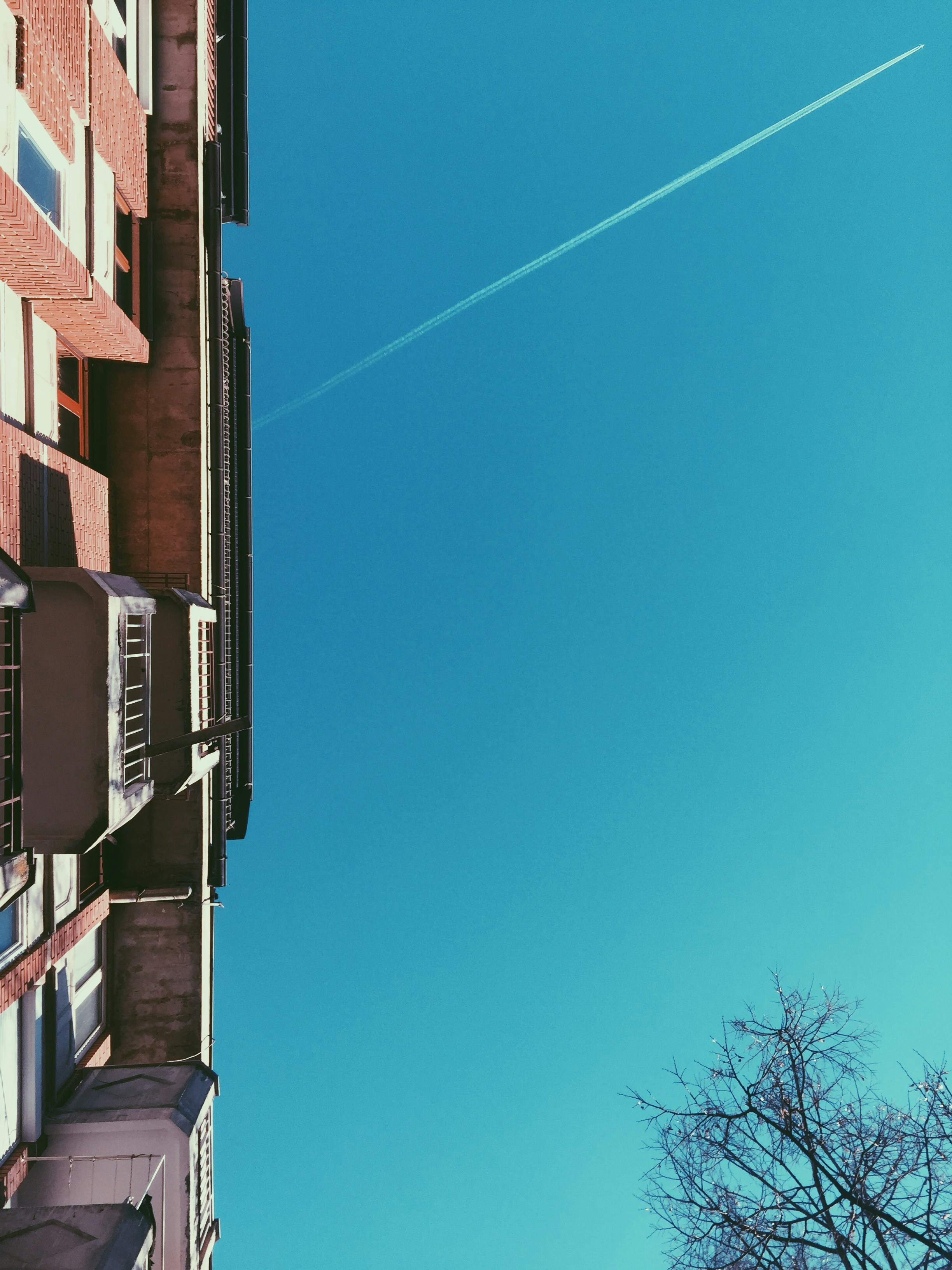 A close-up view of a building's facade reaching towards a clear blue sky, highlighted by a jet's contrail. The image captures the interplay of architecture and atmospheric elements.