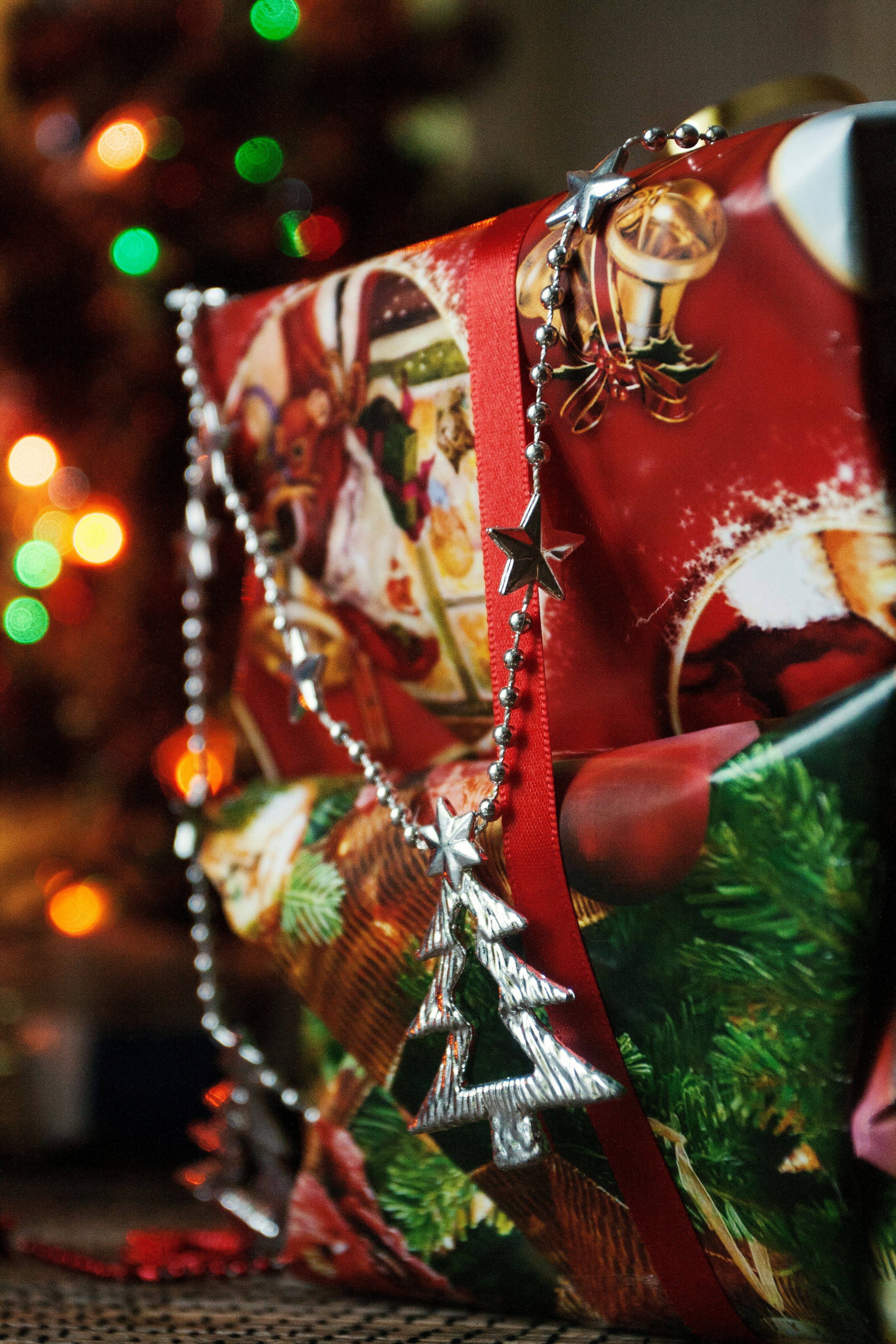 Wrapped Christmas gift in red festive paper features a beaded chain of silver star and tree-shaped charms. Warm holiday lights glow softly as a bokeh backdrop.